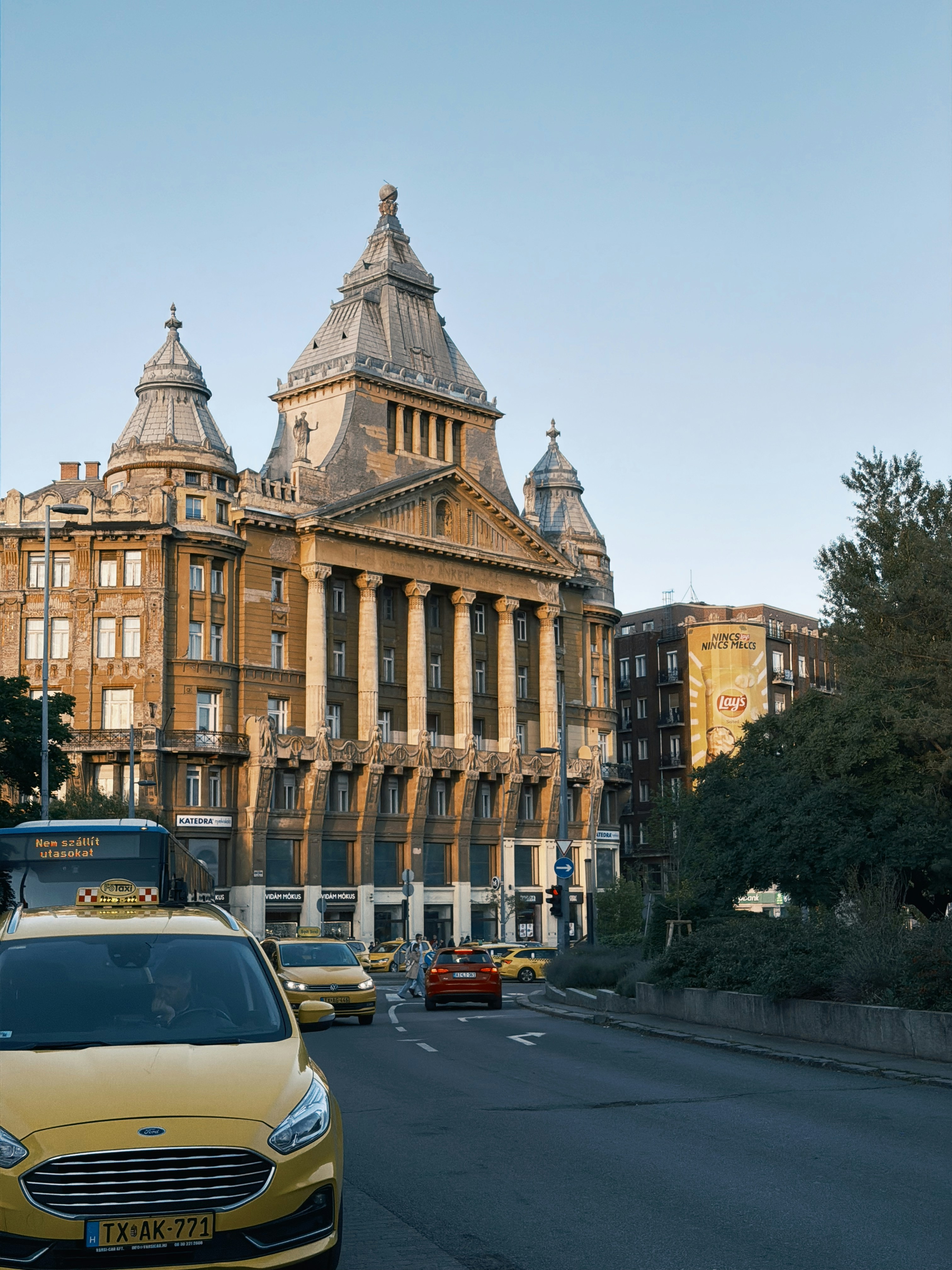 An ornate towered historic building stands behind parked yellow taxis and a Lay's billboard on a city street at golden hour.