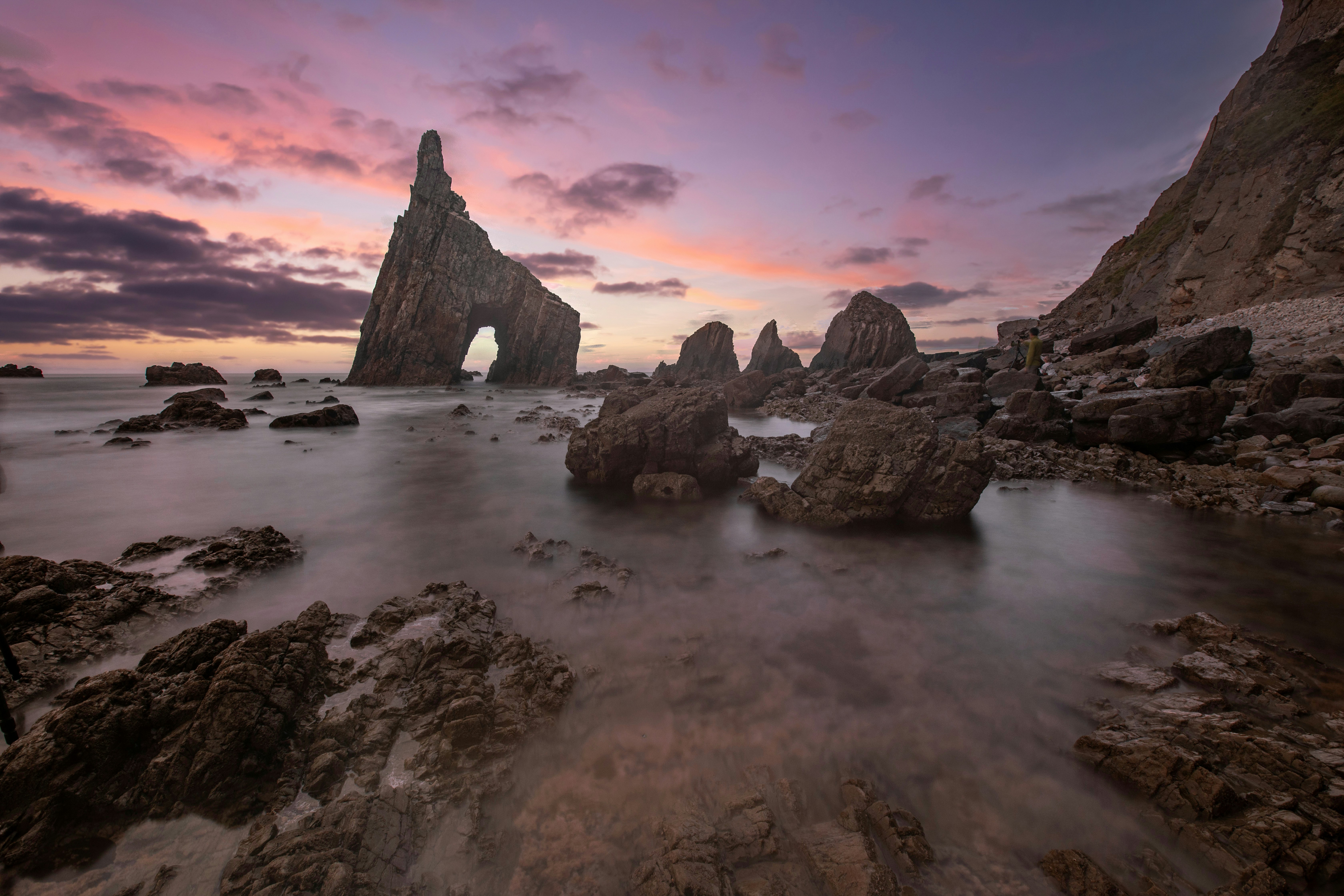 A long exposure shot capturing the epic sunset over Campiecho beach, on the wild Asturian coast of Northern Spain. The iconic natural arch rock formation rises majestically above silky, milky-soft water, smoothed by the exposure time. The sky is painted with dramatic pink, purple, and orange hues, as clouds stretch into the twilight. Two tiny photographers are visible on the rocky shore to the right, offering a sense of scale to nature's vastness.