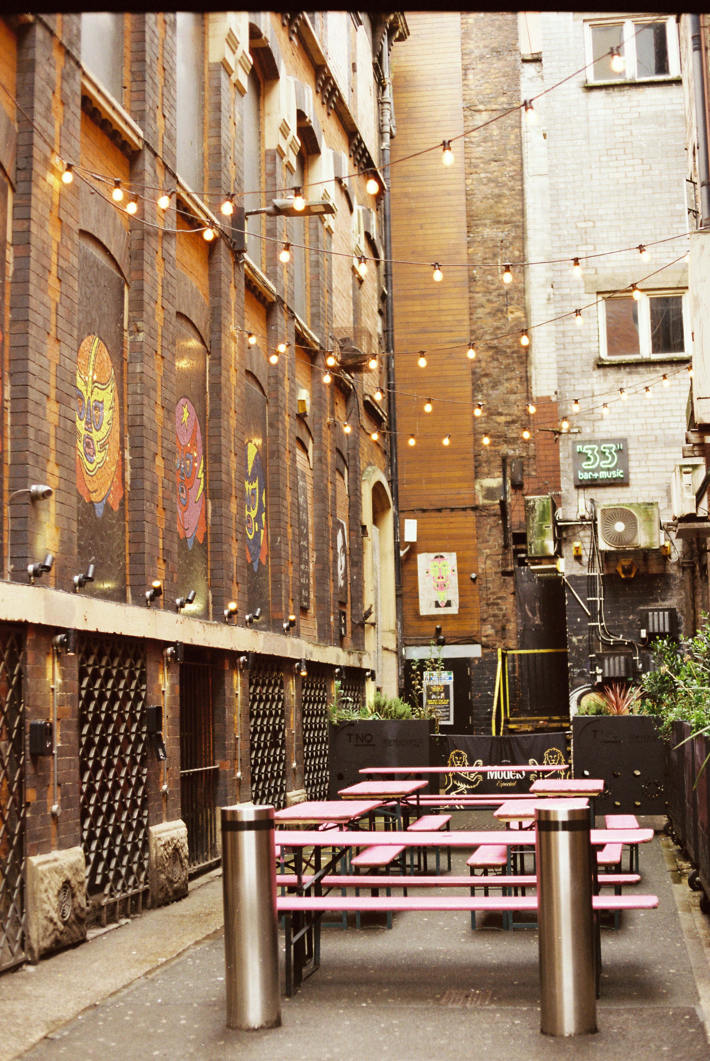 Pink picnic benches sit in a courtyard strung with festoon lights and lined with painted murals and a bar sign.