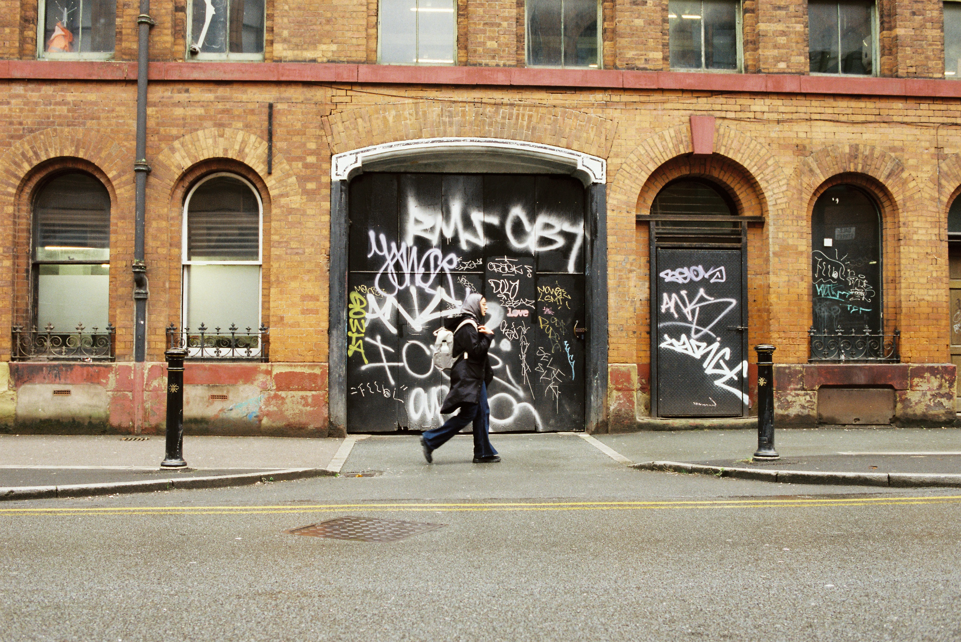 A figure with a backpack walks past a heavily tagged black loading door set in a worn brick warehouse facade.