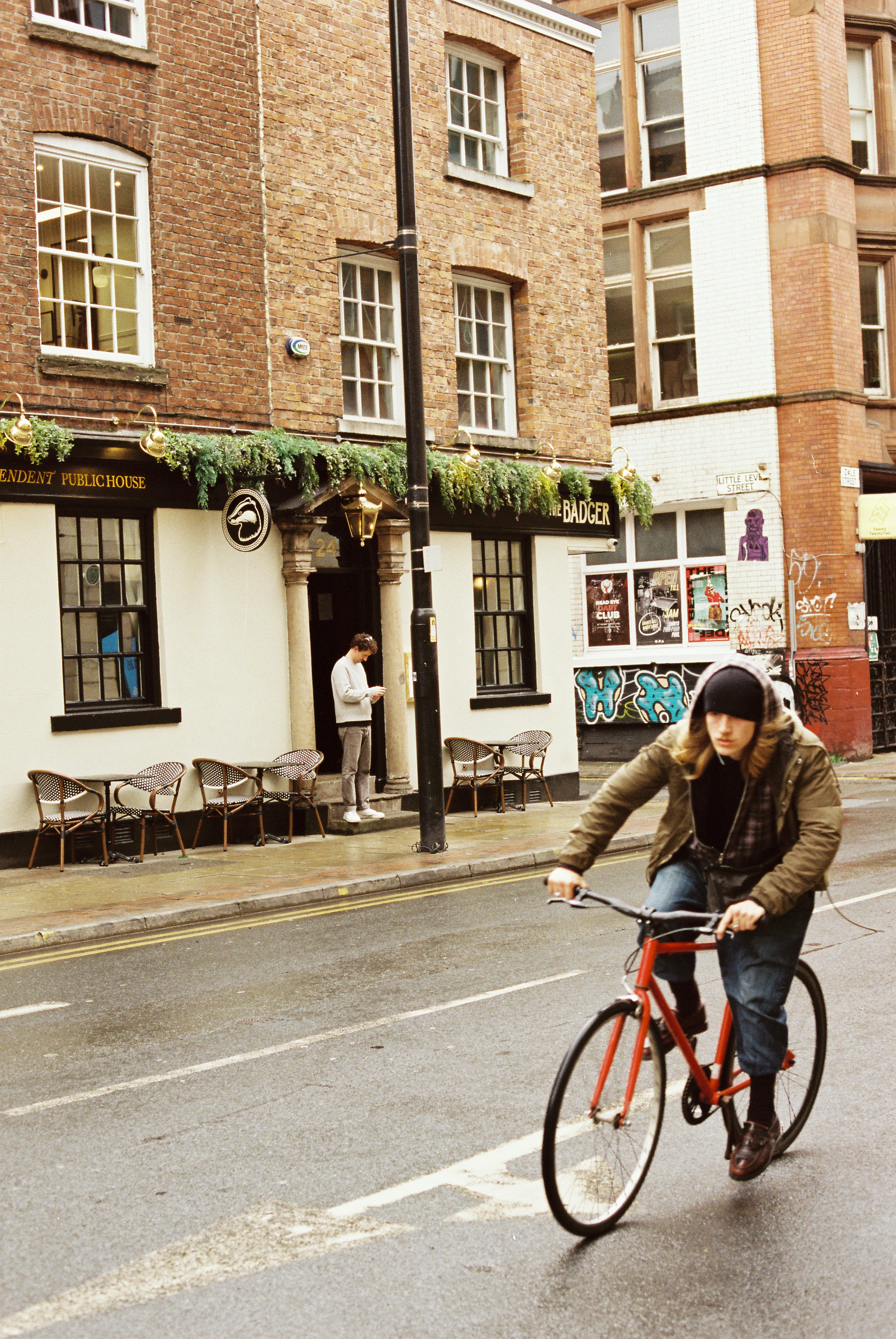 A man cycles past The Badger pub on a damp street while another stands reading outside under hanging greenery.