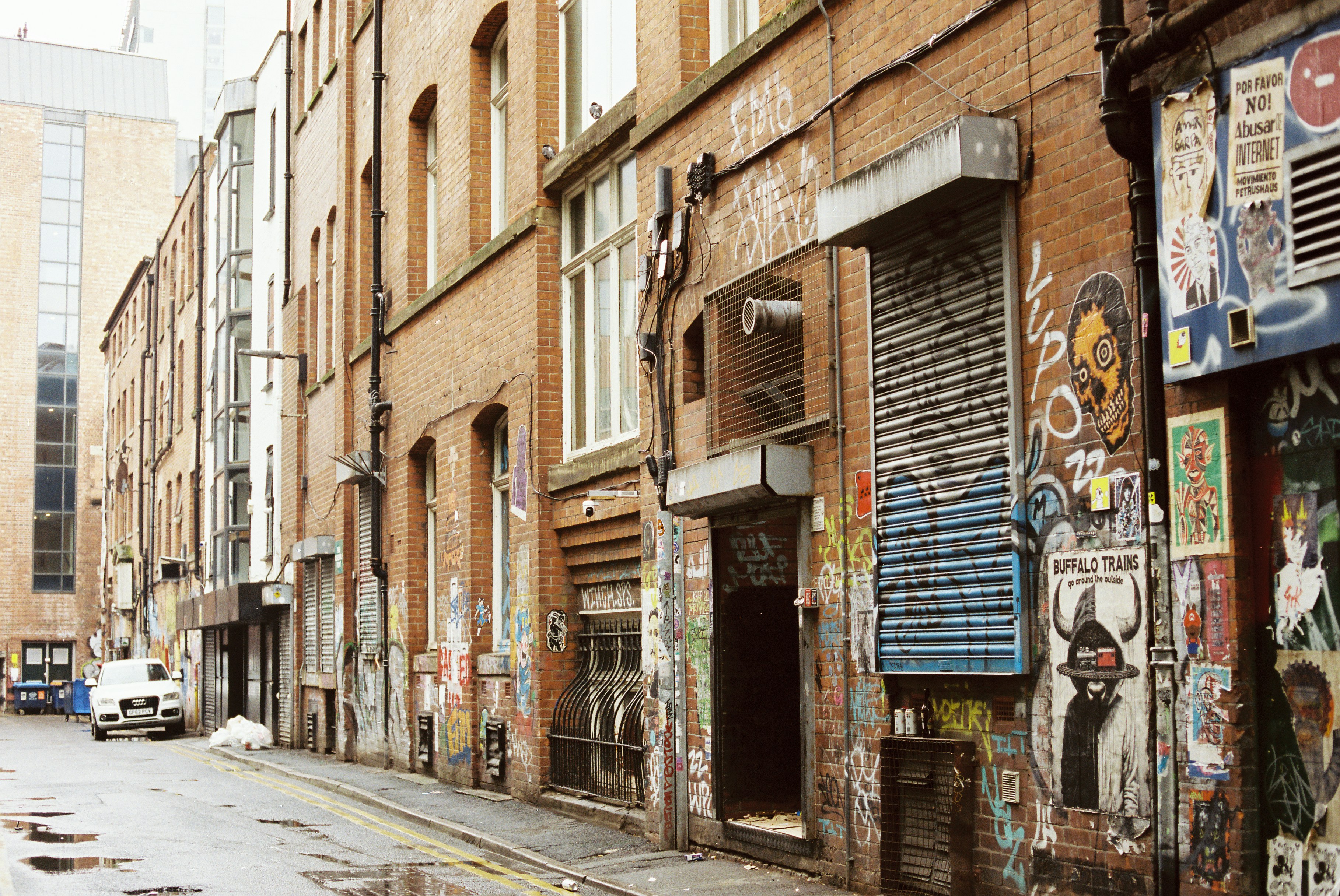 A graffiti and paste-up covered brick backstreet stretches away with a white car parked along the kerb.