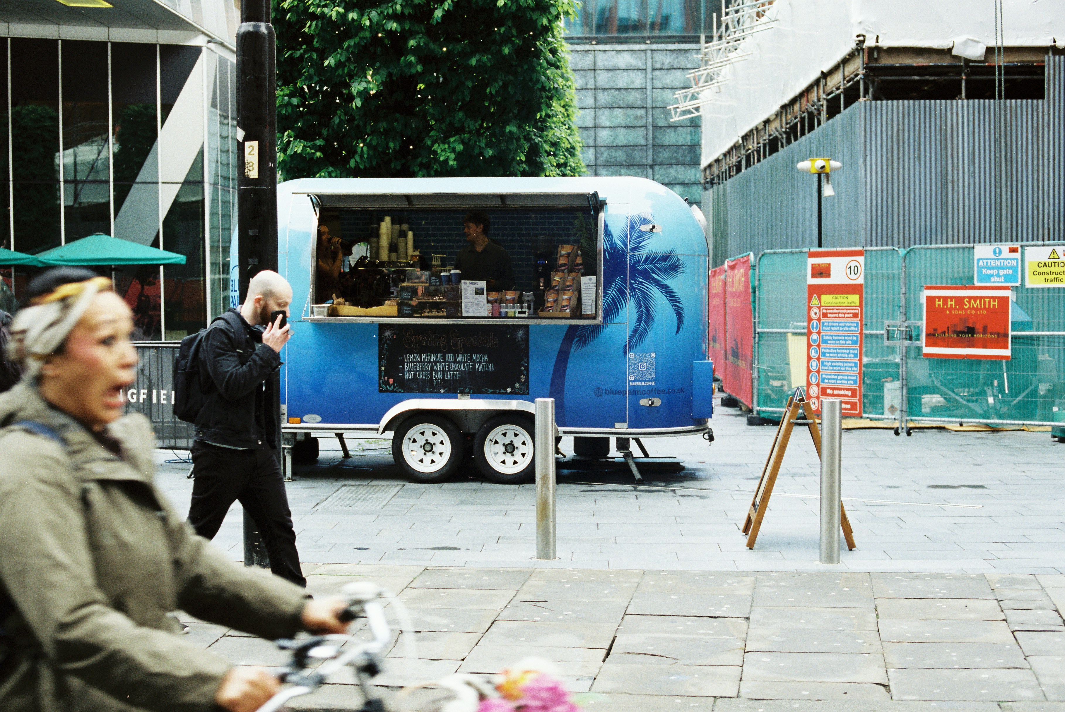 A vintage blue coffee trailer sits in a plaza as a man on a phone and a woman with a bike pass in front.