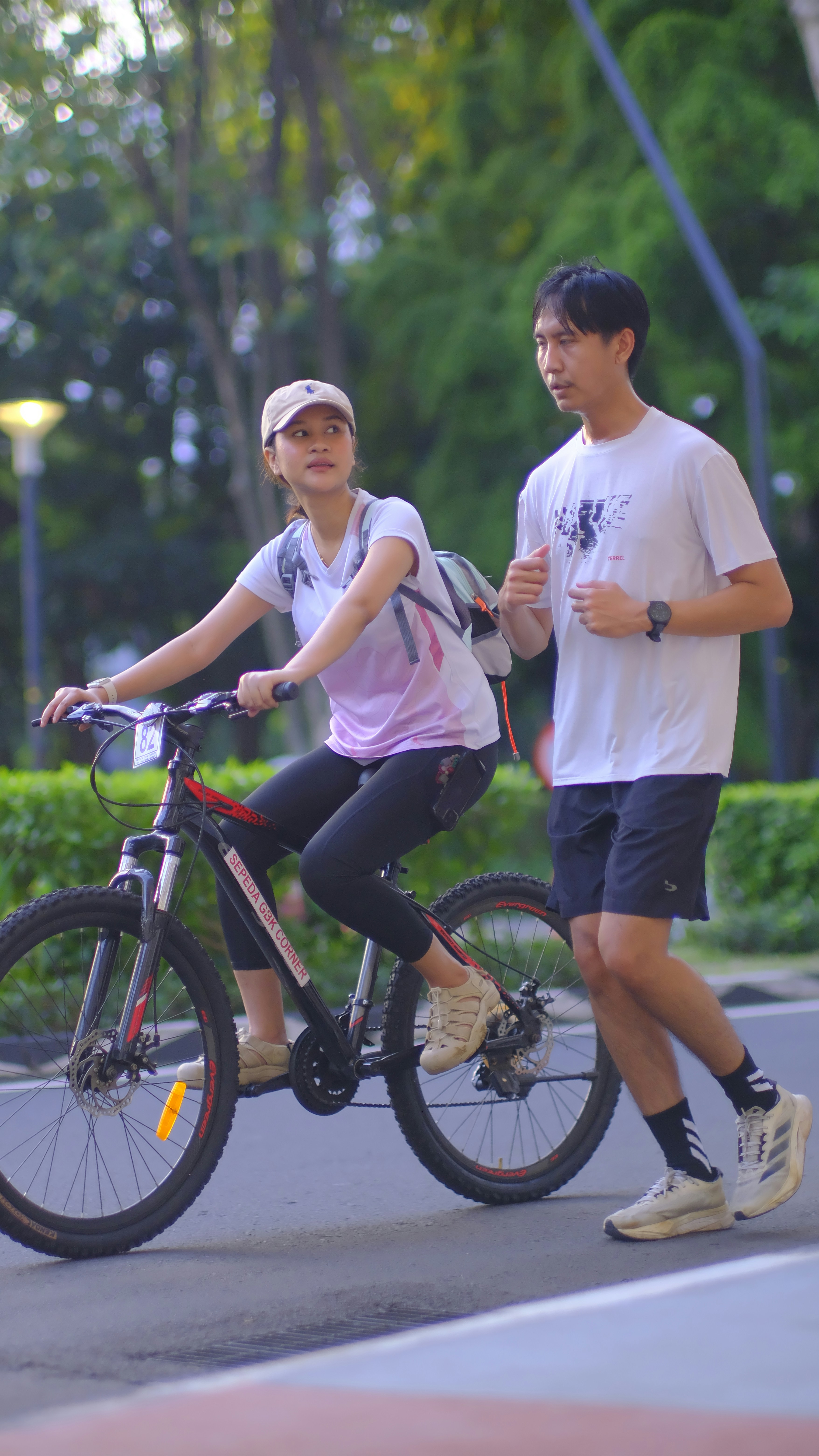 Two people in a park: a woman riding a bike while a man walks beside her, both engaged in a casual outdoor scene.