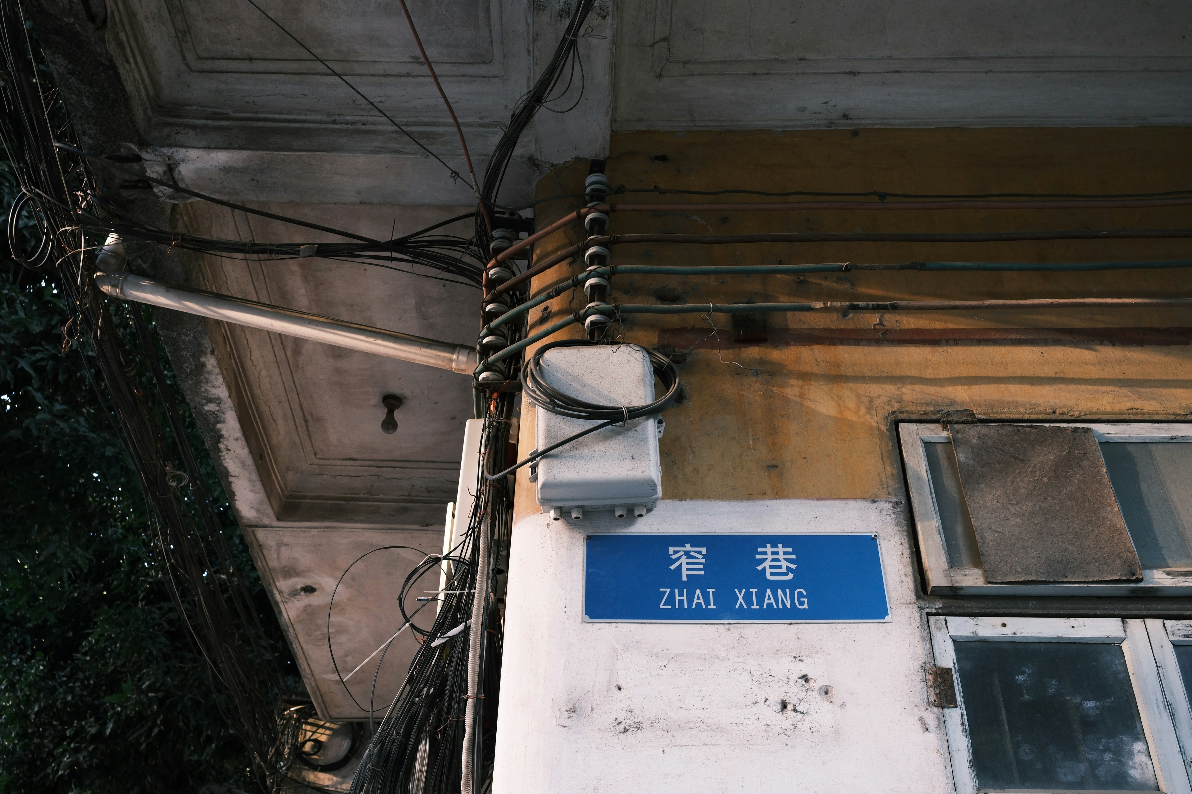 Blue and white street sign reading ZHAI XIANG on a weathered building facade with tangled cables and a window nearby.