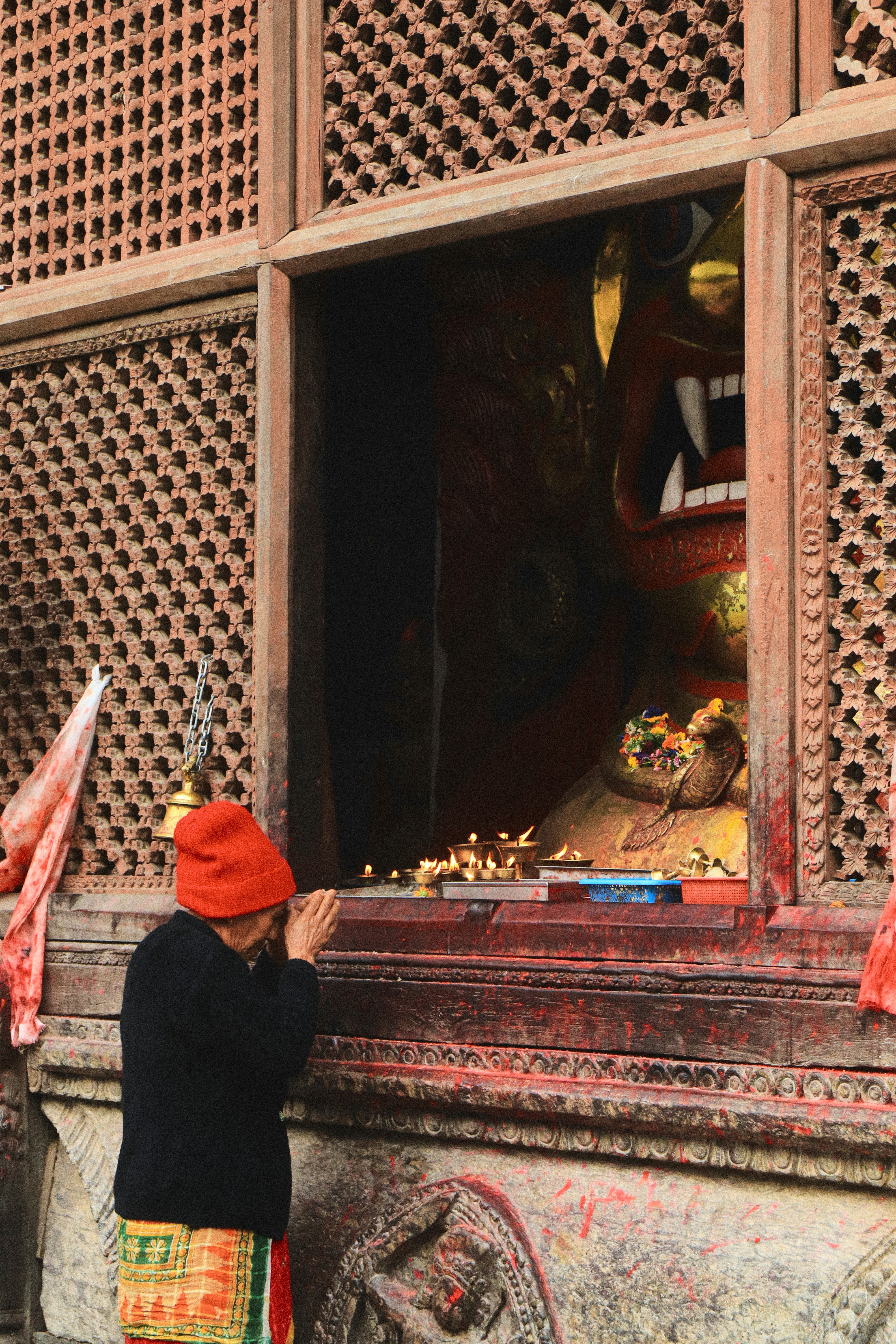 Devotion at the Gate: A Worshipper in Red Knit and Orange Sari Foreground a Fierce Masked Guardian