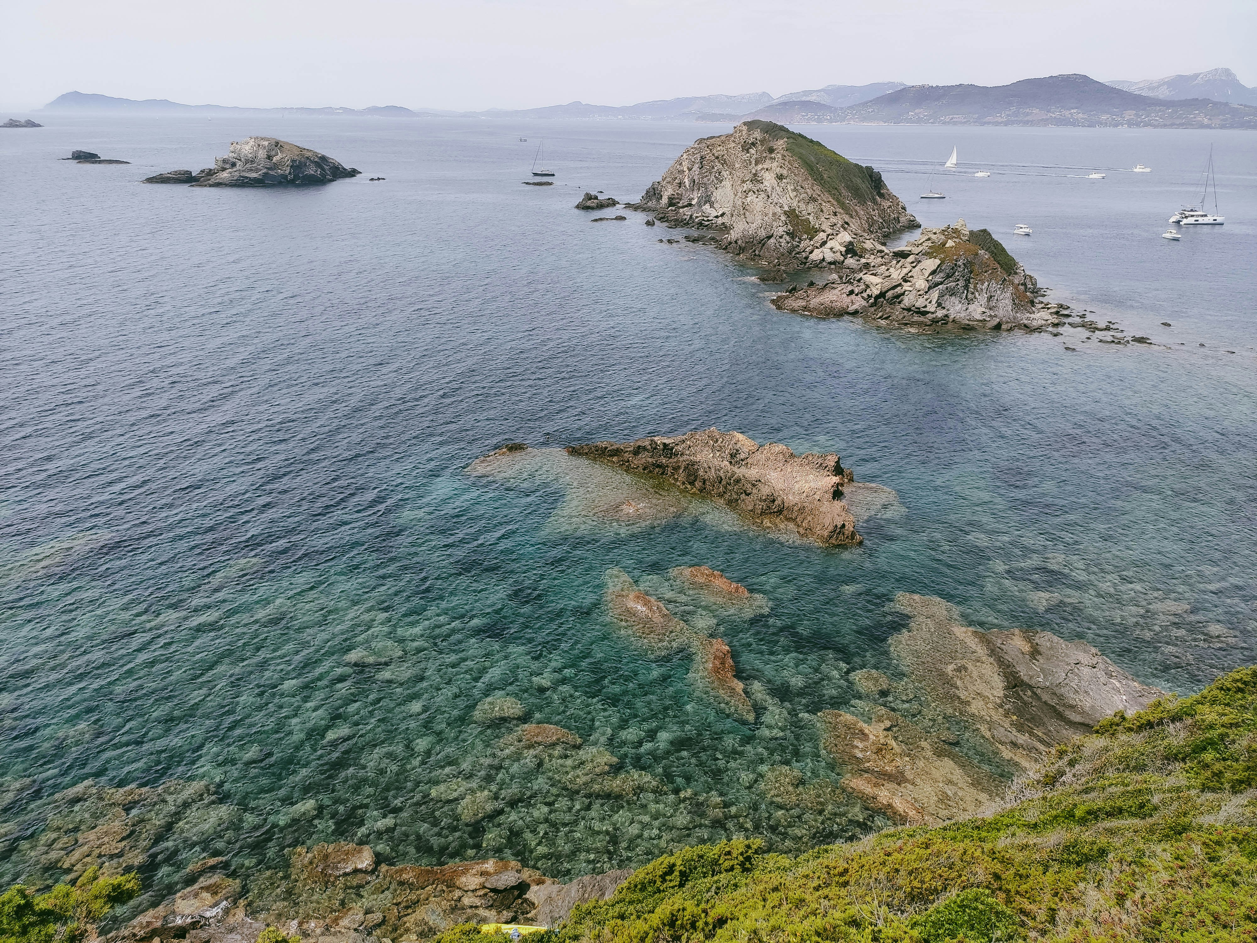 Clear turquoise water washes over submerged rocks near a rocky outcrop; distant boats dot the horizon and a hazy coastline forms the backdrop. This is a landscape scene with natural textures and color.