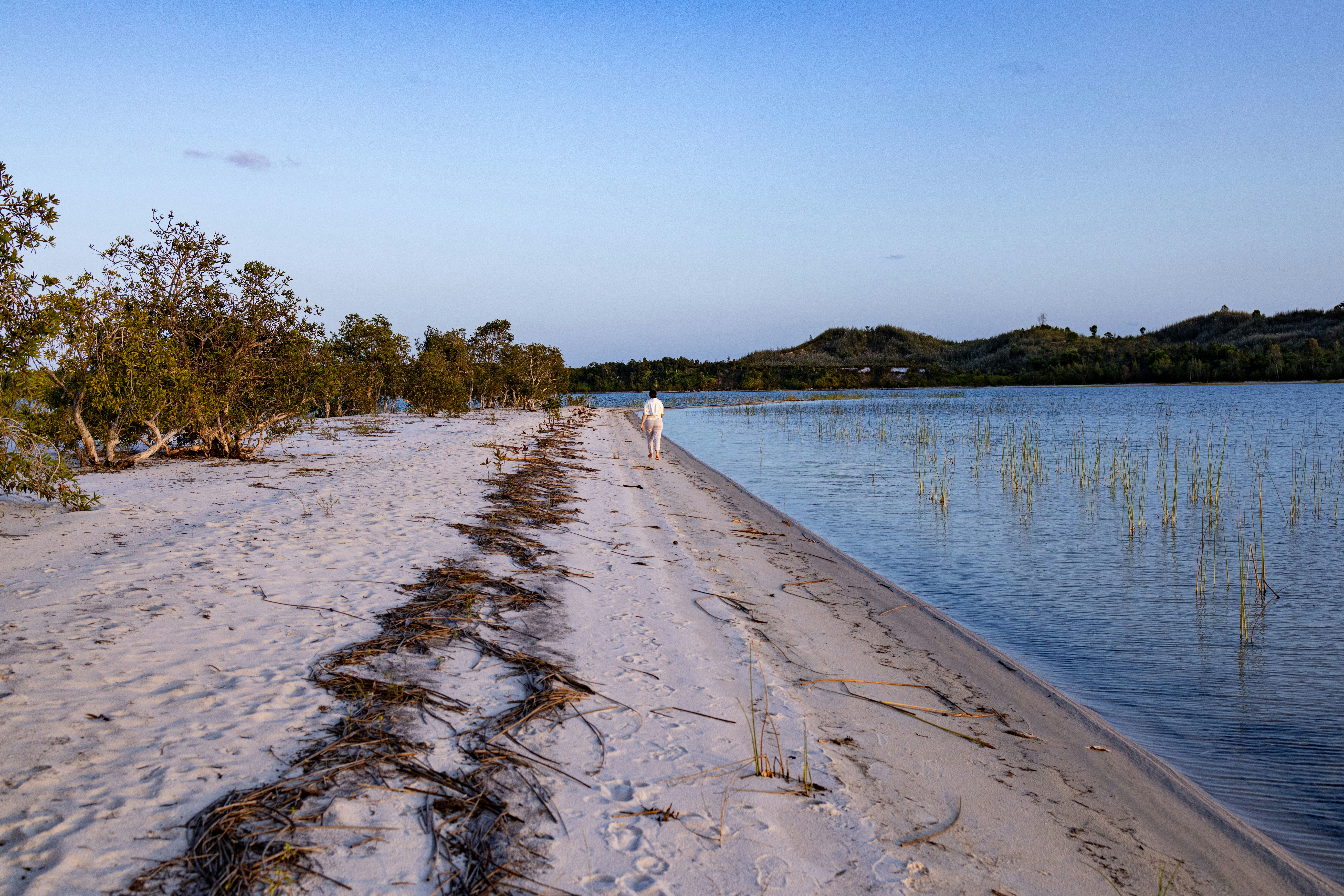 A person walks along a sandy path beside calm water.