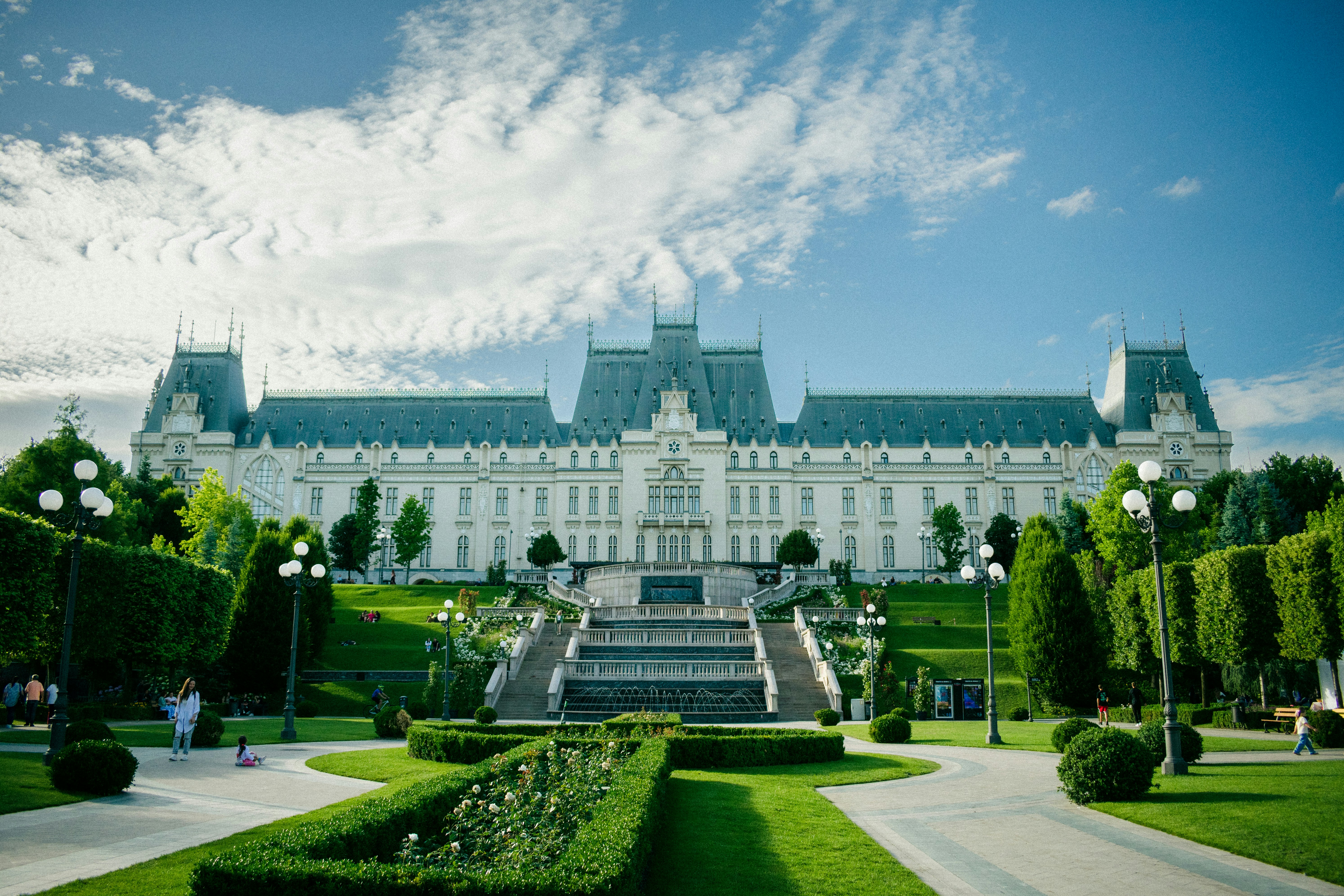 Grand white palace with layered terraces and manicured gardens under a blue sky with scattered clouds.