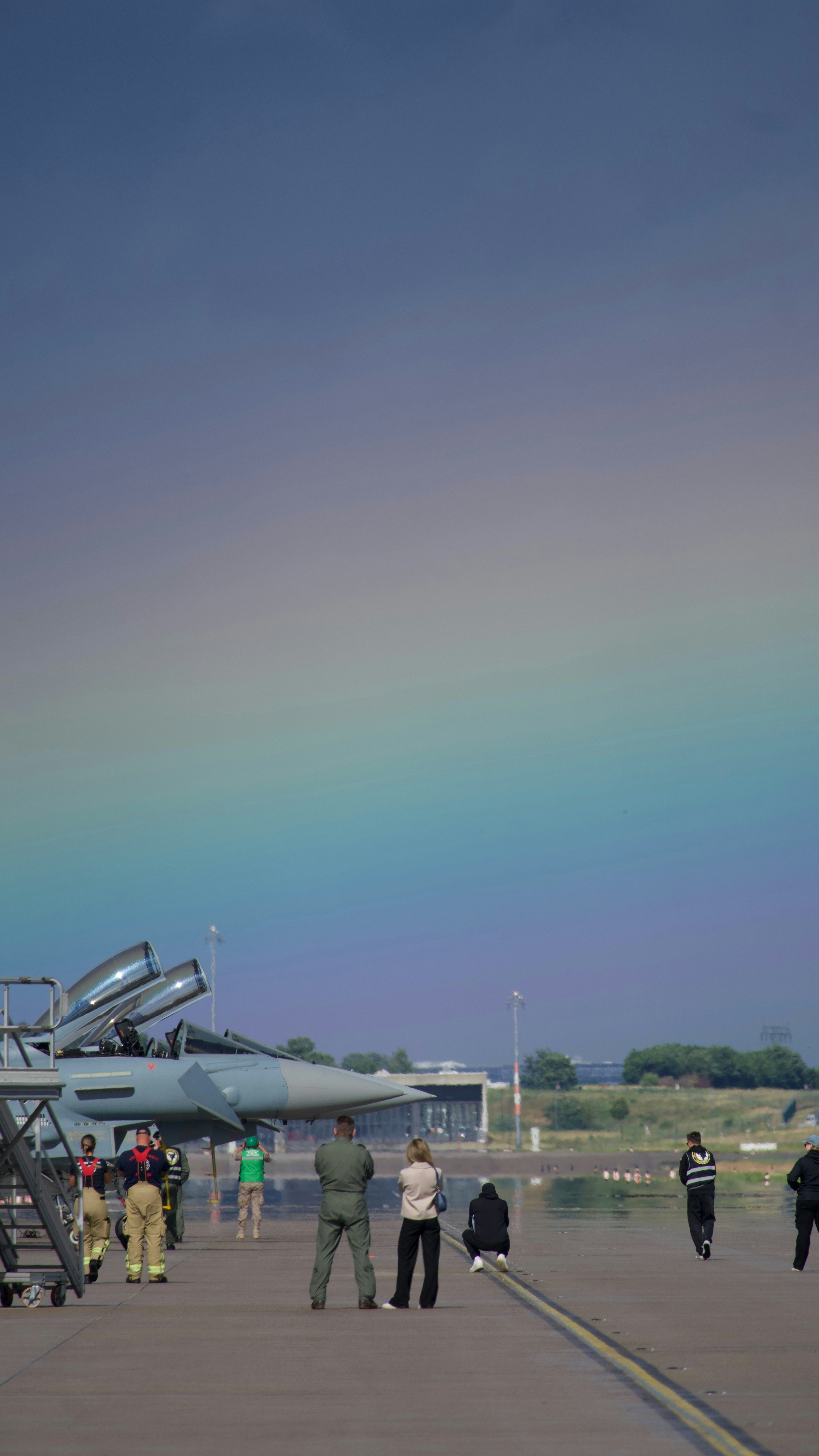 Rainbow over a military ramp: figures and jet under a pastel sky