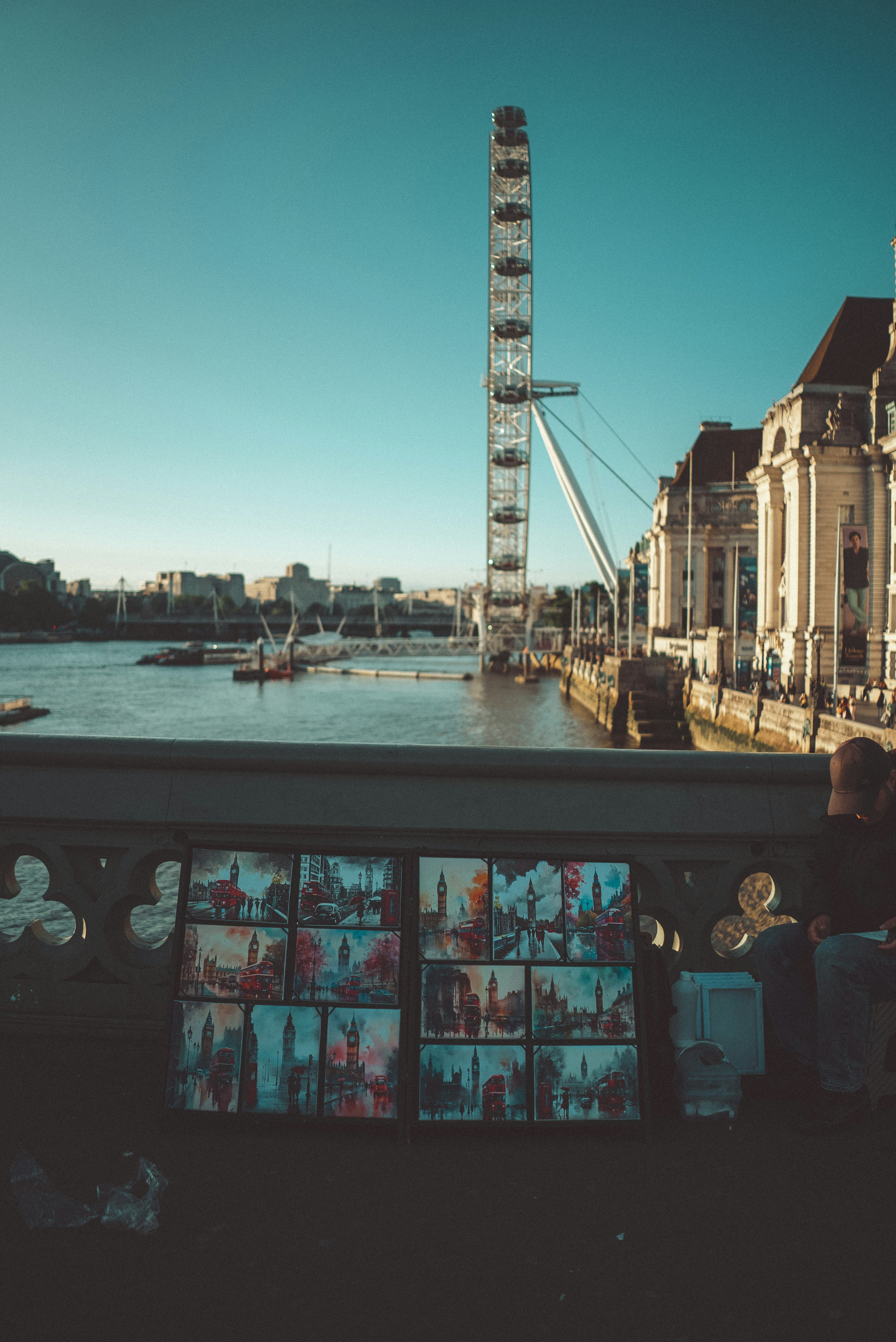 Colorful prints for sale line a railing beside the Thames with the London Eye visible in the background on a clear day.