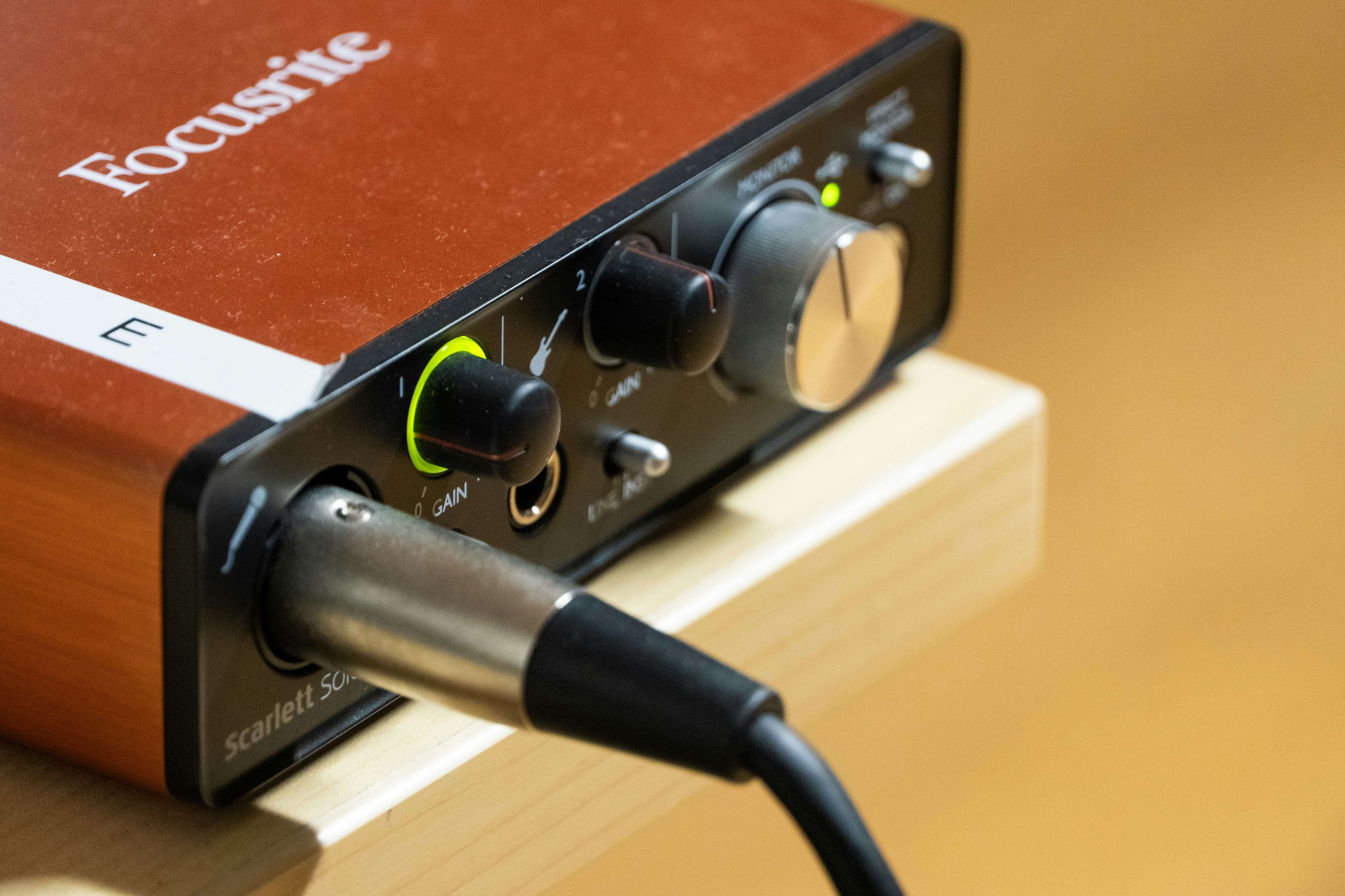Close-up of a Focusrite audio interface with visible front panel knobs and a plugged-in cable, emphasizing tactile controls and warm wood backdrop.