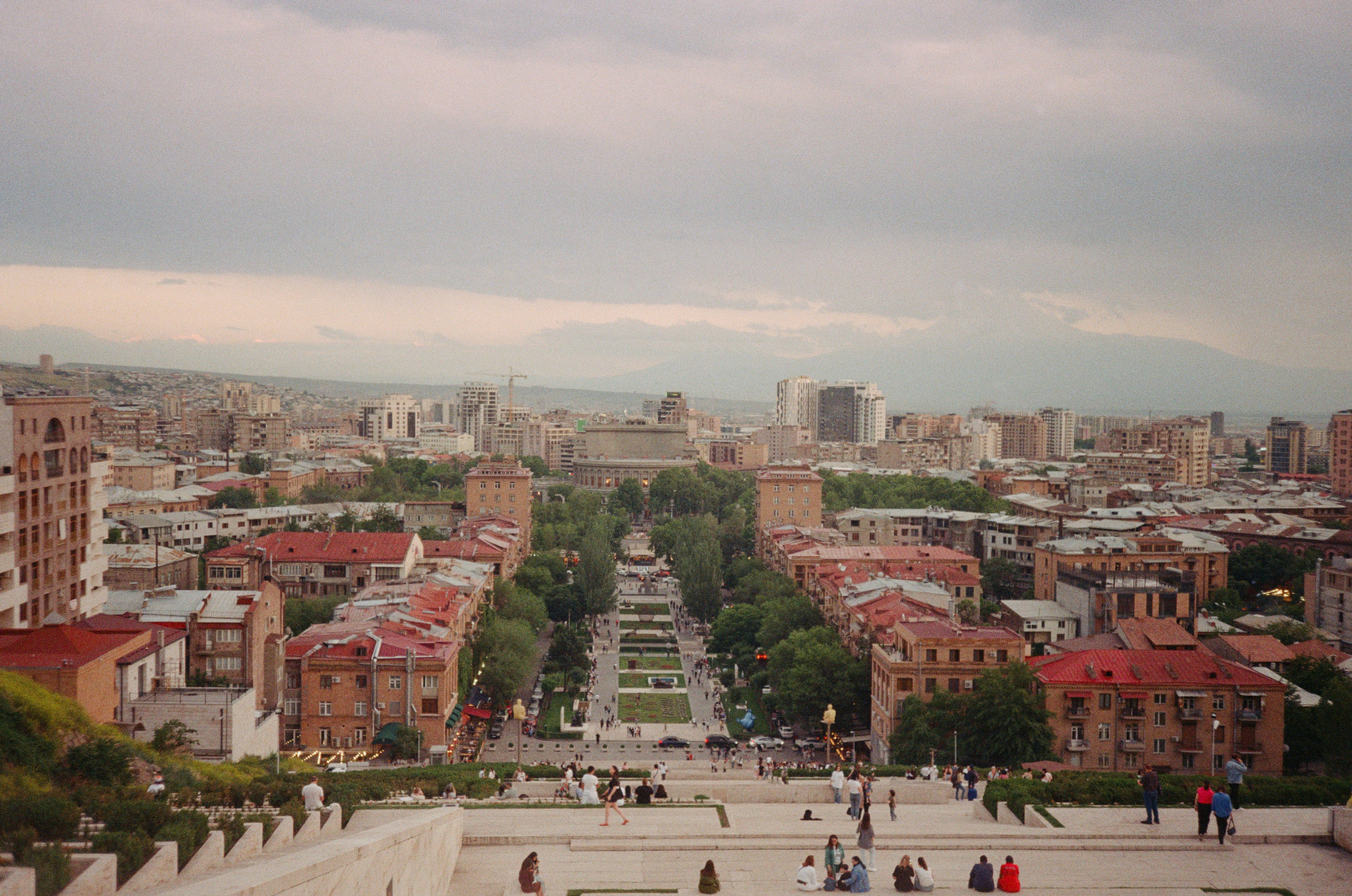 A view over Yerevan, Armenia from the Cascade, with city buildings, red rooftops, mountains, and cloudy sky. 35mm film photo shot on an Olympus Mju-1.