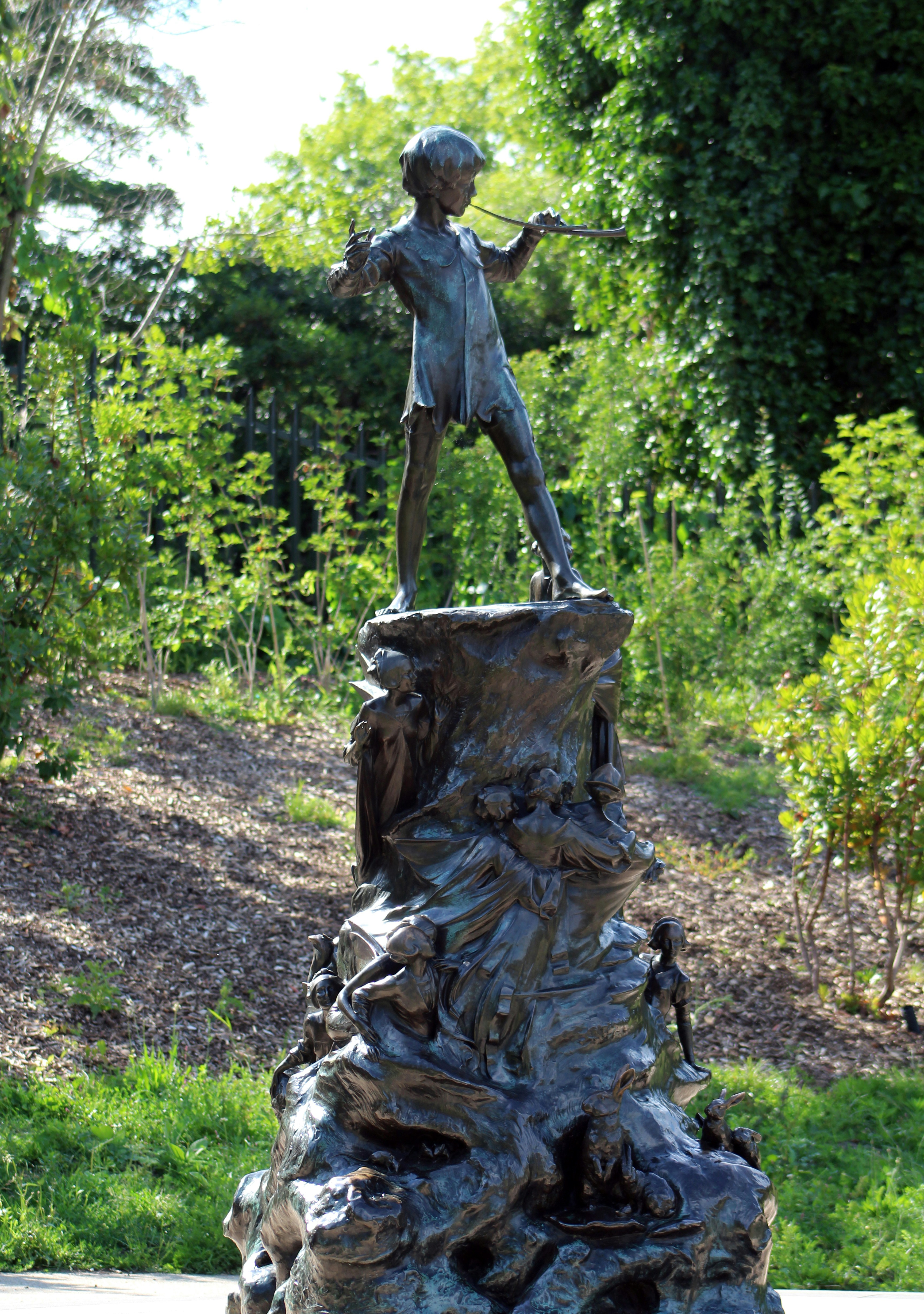 Bronze statue of a boy atop a rough pedestal, arm extended with a rifle, set against lush greenery in a park. A sculpture-focused outdoor scene with natural light and textured pedestal details.