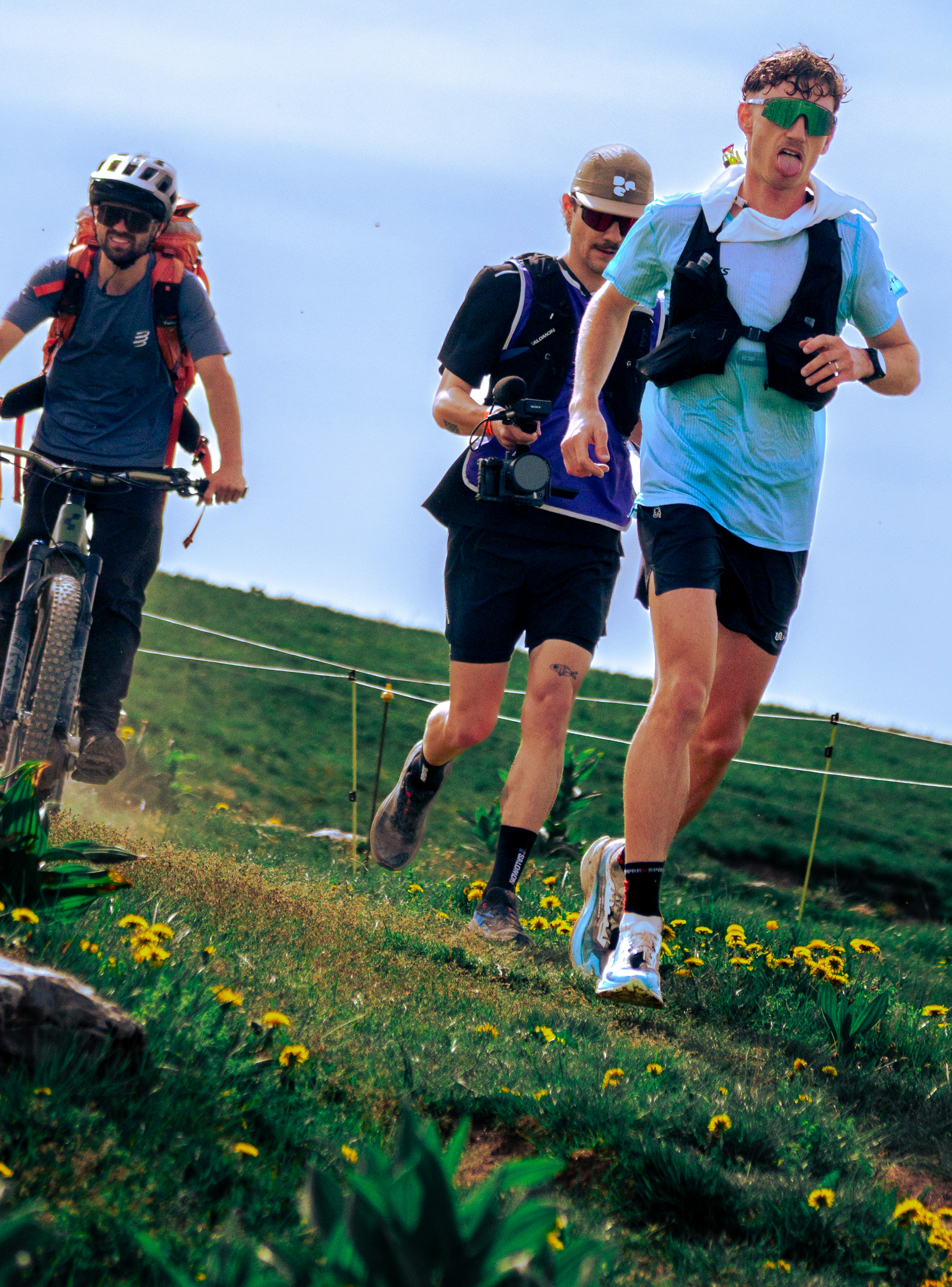Three runners sprint along a grassy hillside with wildflowers, wearing hydration vests and gear. The scene captures motion and outdoor adventure, with a vivid blue sky serving as backdrop.