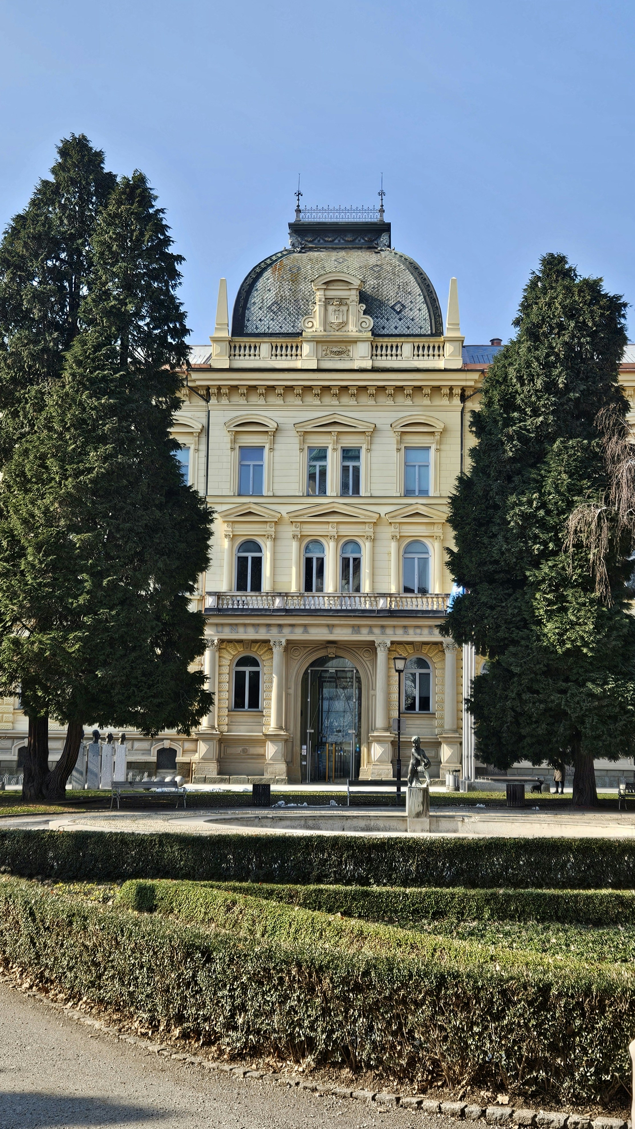 Elegant historic building framed by two tall trees with a sculpted fountain in the foreground; architectural detail and a clear blue sky define the scene.