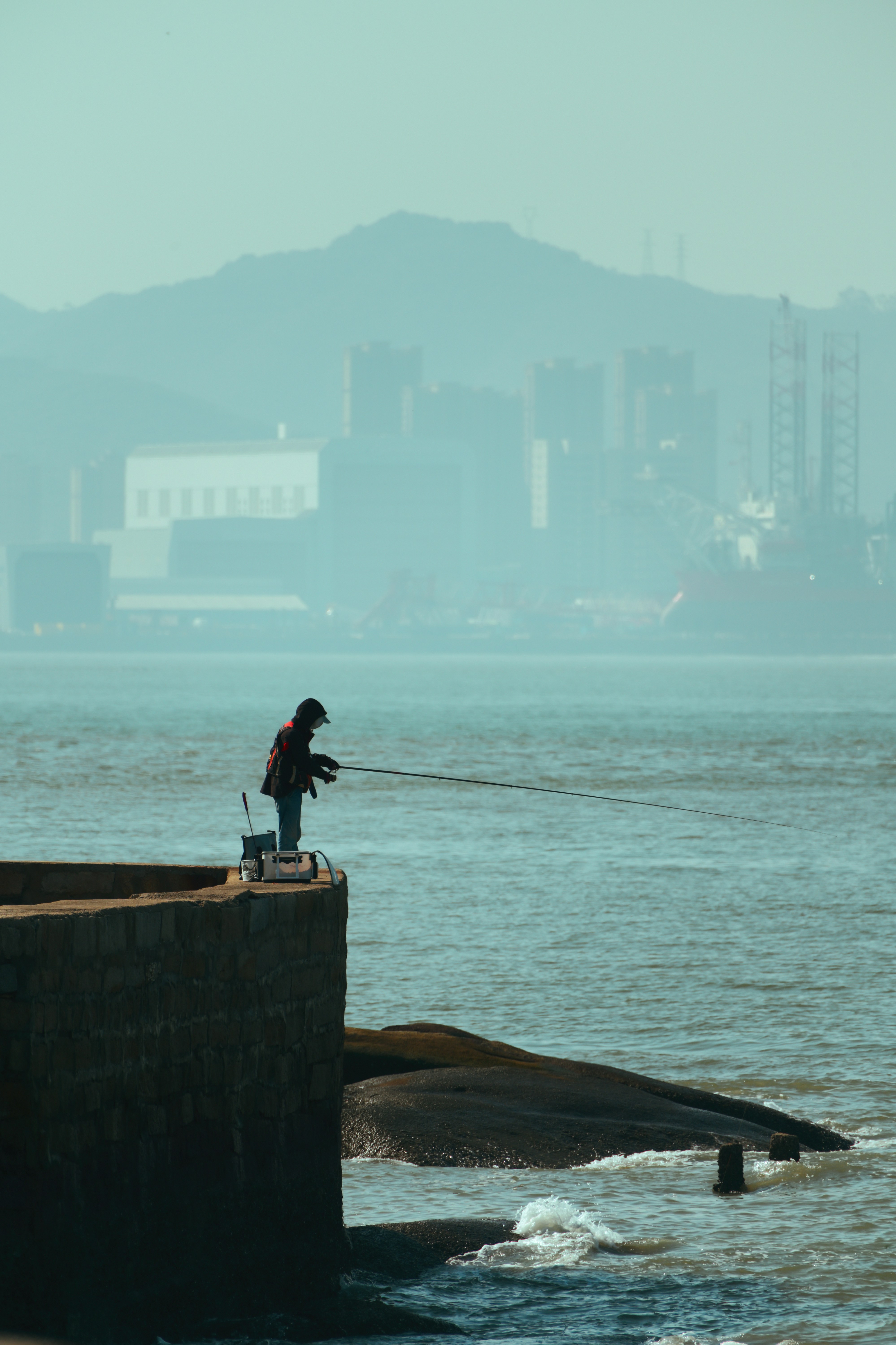 Solitary Angler at the Edge: A Quiet Pier Against a Distant Industrial Landscape