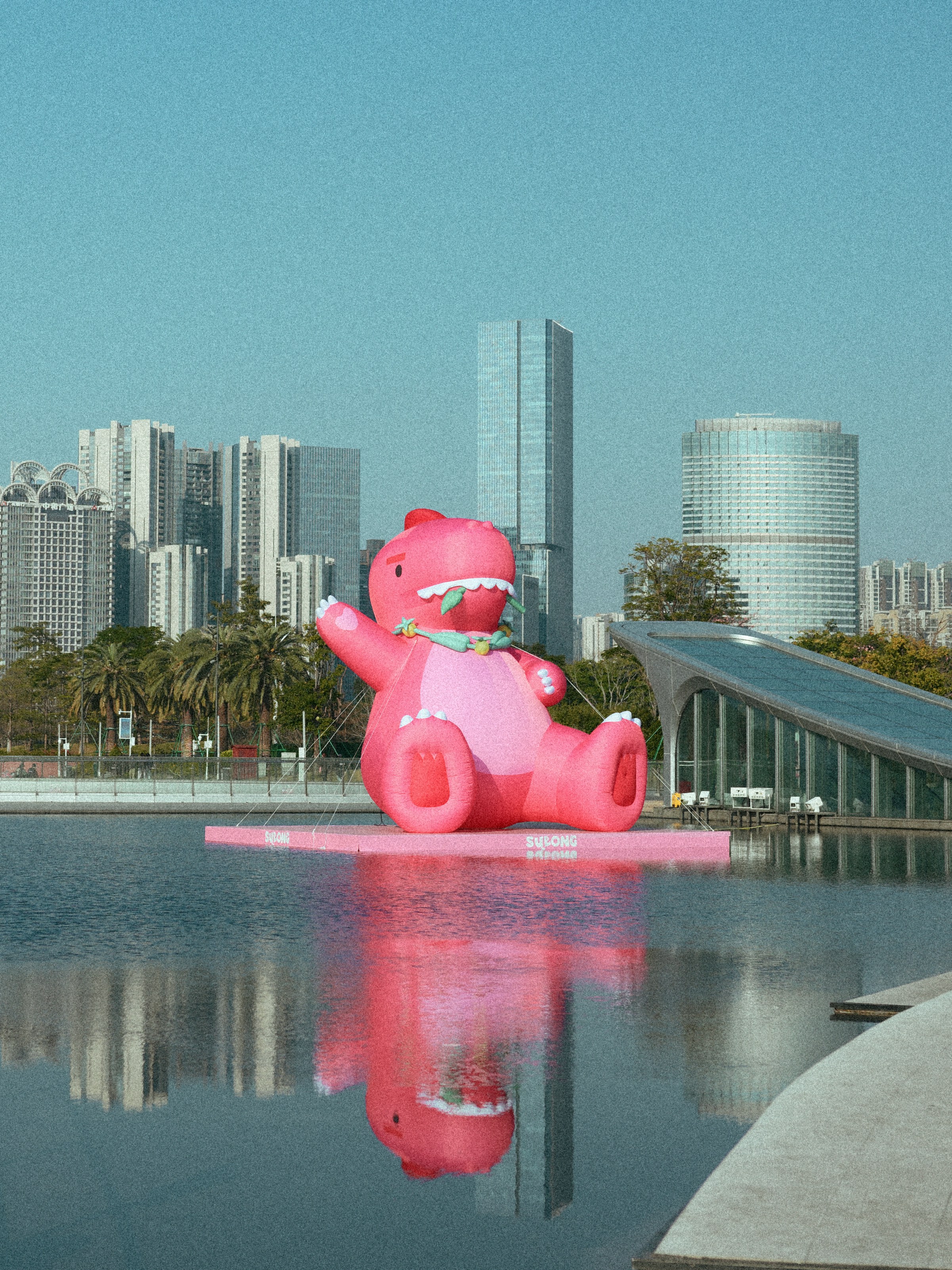Pink inflatable dinosaur floats on water before a city skyline.