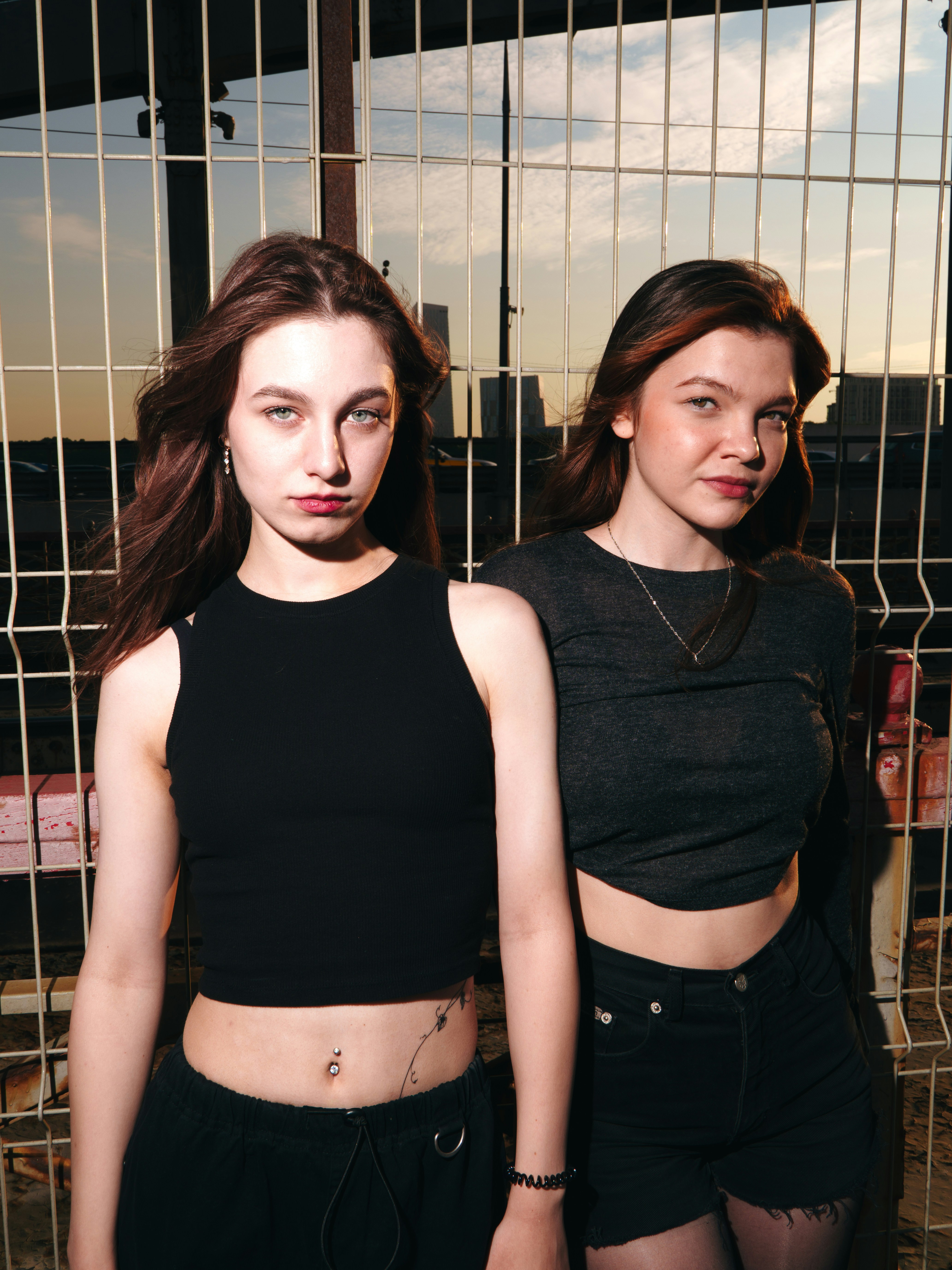 Two young women stand in front of a metal fence.