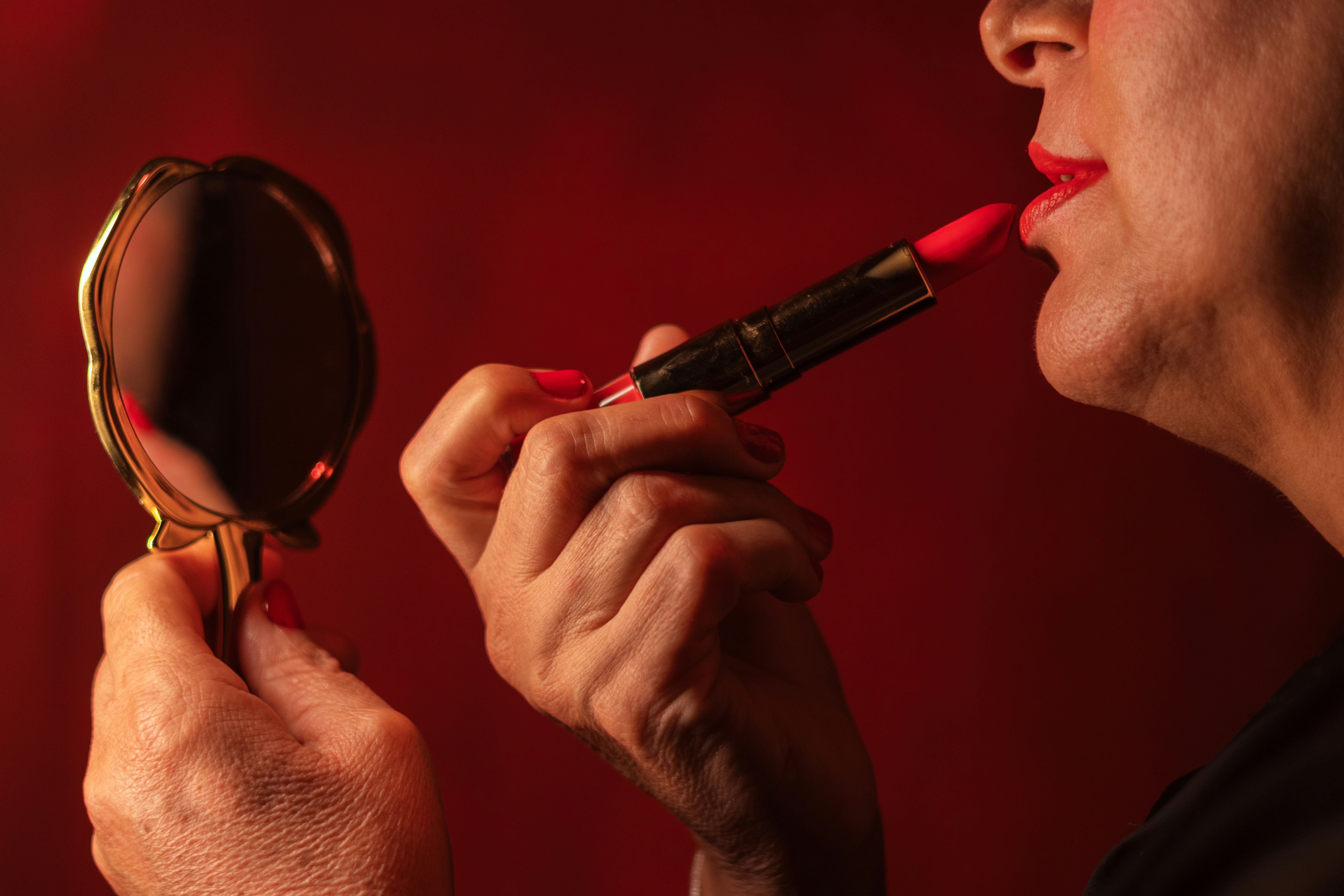 Close-up of a person applying red lipstick while holding a small mirror against a deep red backdrop.