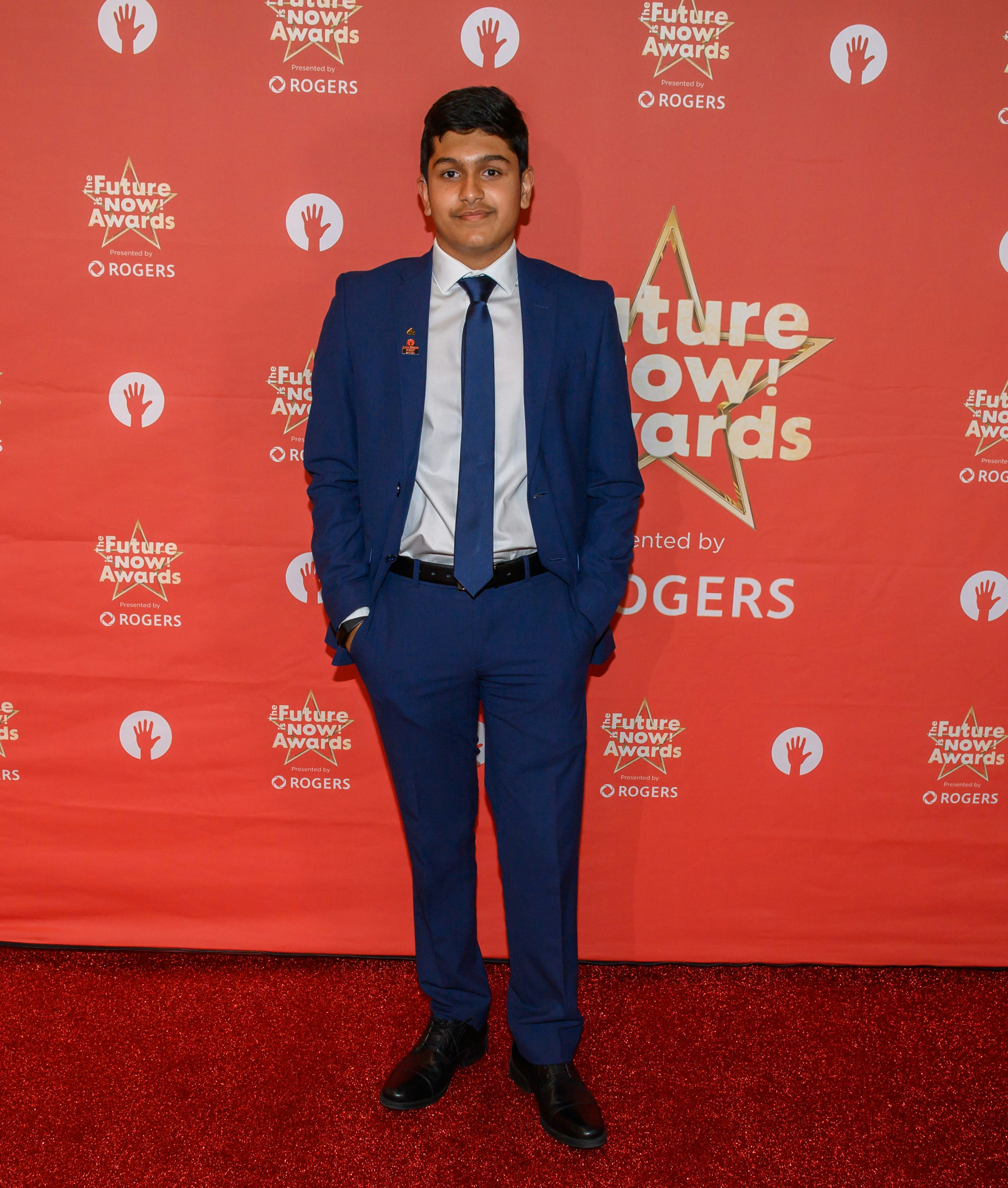 Young man in a tailored blue suit on a red carpet backdrop with Future Now Awards branding. A polished formal portrait highlighting the subject against a branded environment.