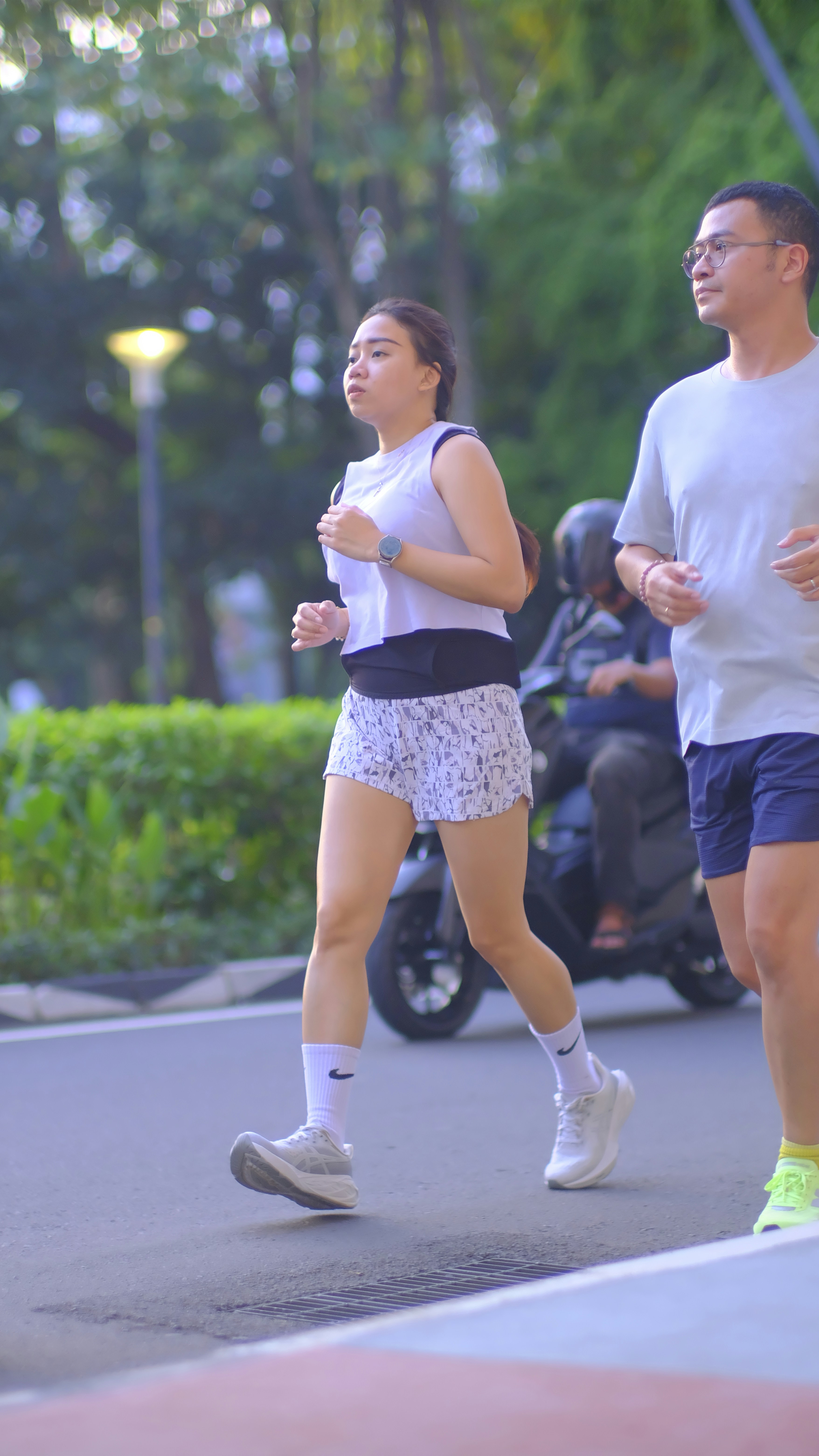 Two joggers move along a city park road with trees and greenery in the background; a woman leads slightly while a man runs beside. This is a candid street shot with natural motion and soft color tones.