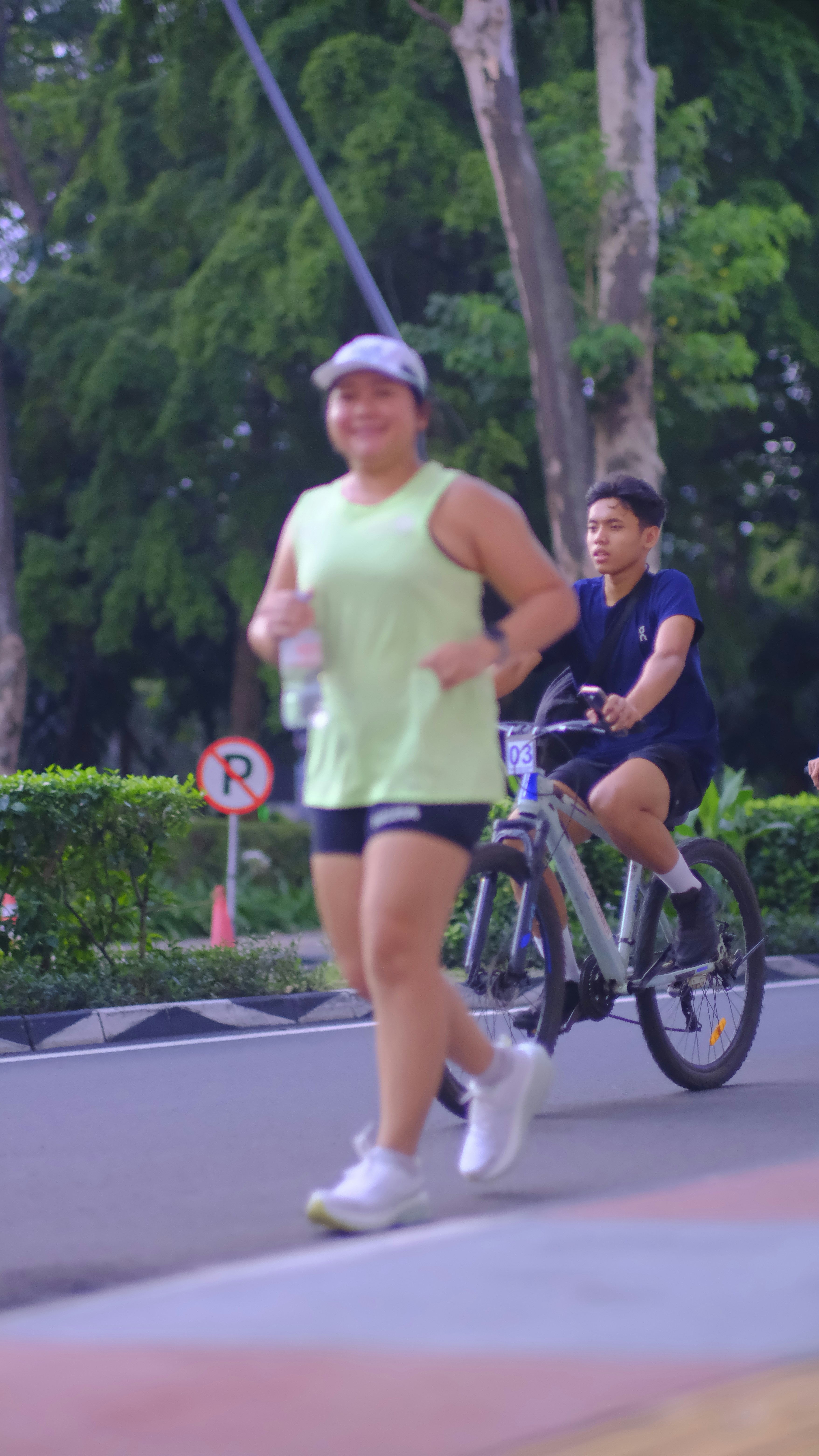 A blurred runner in neon green top crosses in front of a cyclist on a paved park path, with trees and signage in the background. The scene emphasizes motion and urban leisure.