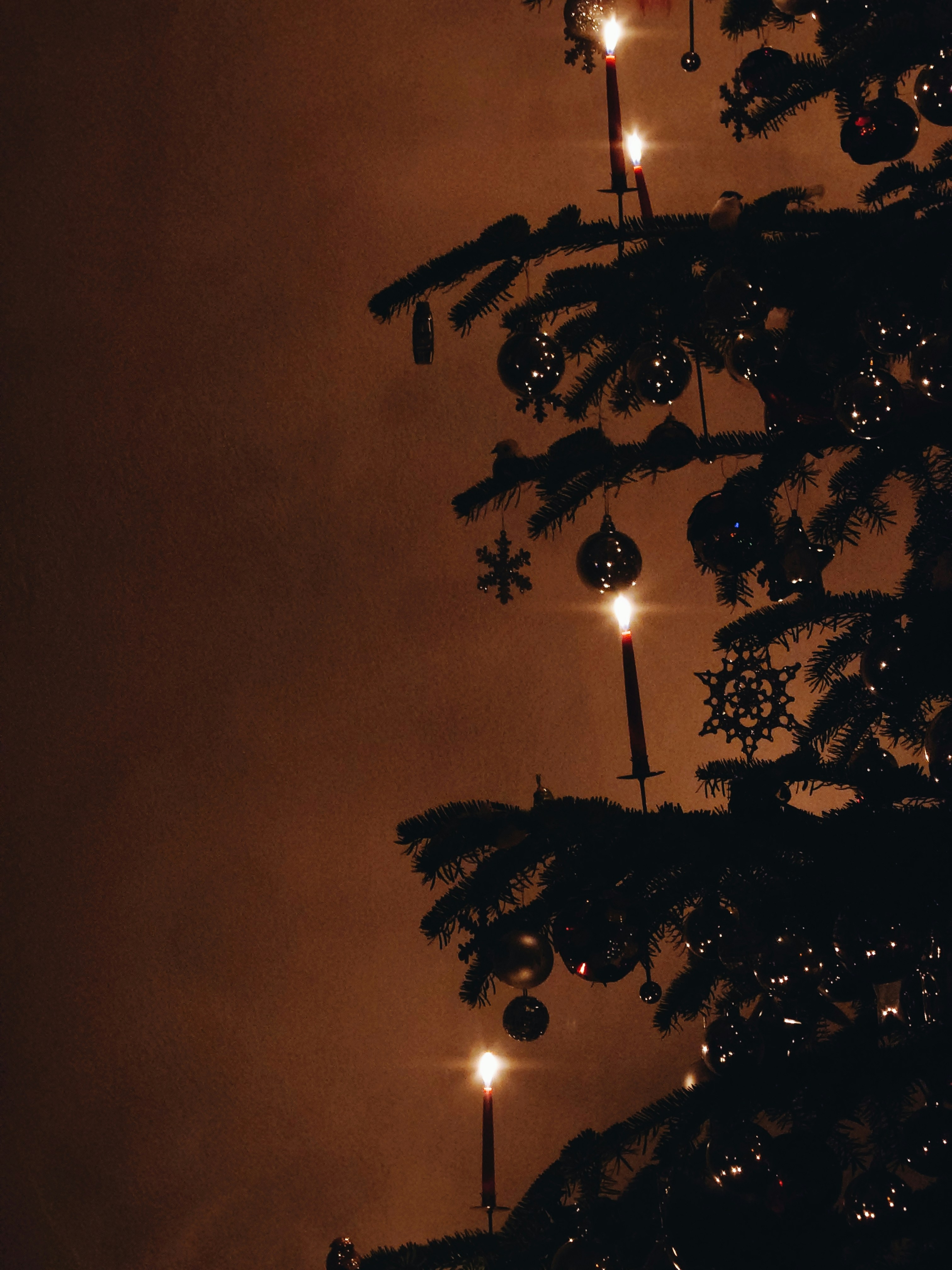 A warm, candle-lit Christmas tree silhouette with hanging ornaments and candles illuminating dark surroundings. The scene emphasizes festive decor and low-light ambiance.