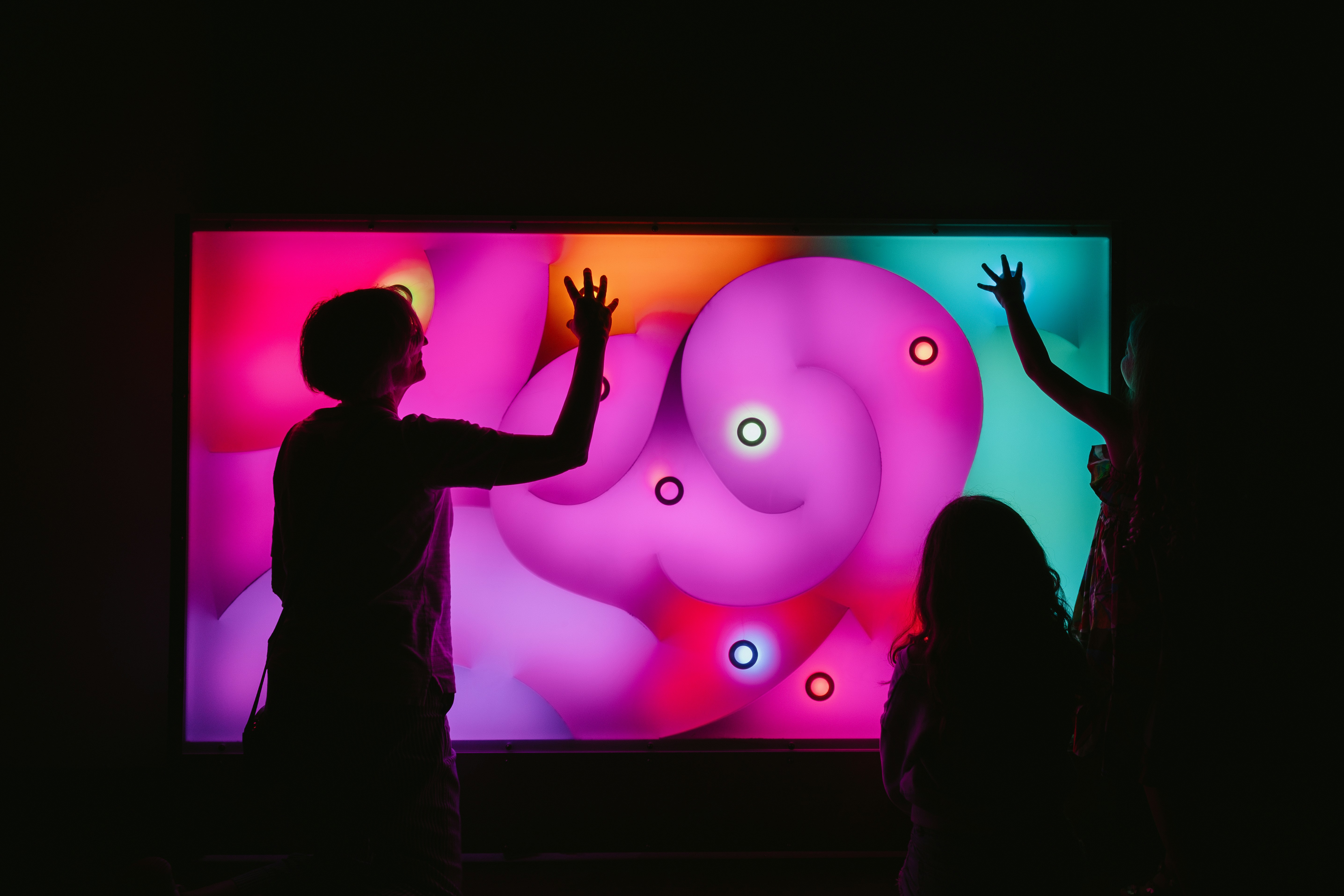 Silhouettes interact with a vibrant, glowing interactive screen in darkness.