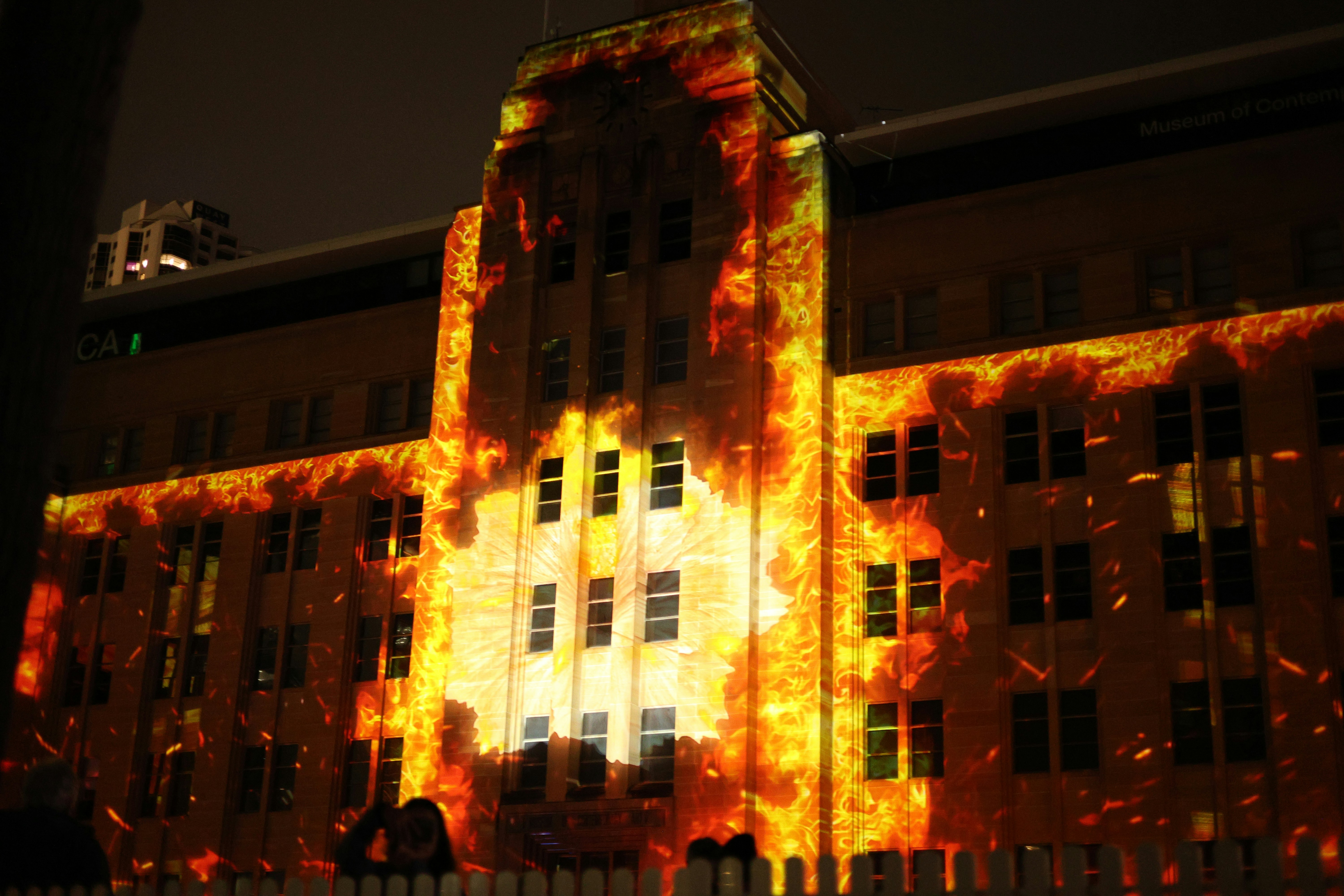 Night-lit building façade bursts with fiery projections, casting dramatic orange and yellow glows across windows and silhouettes of onlookers at the base.