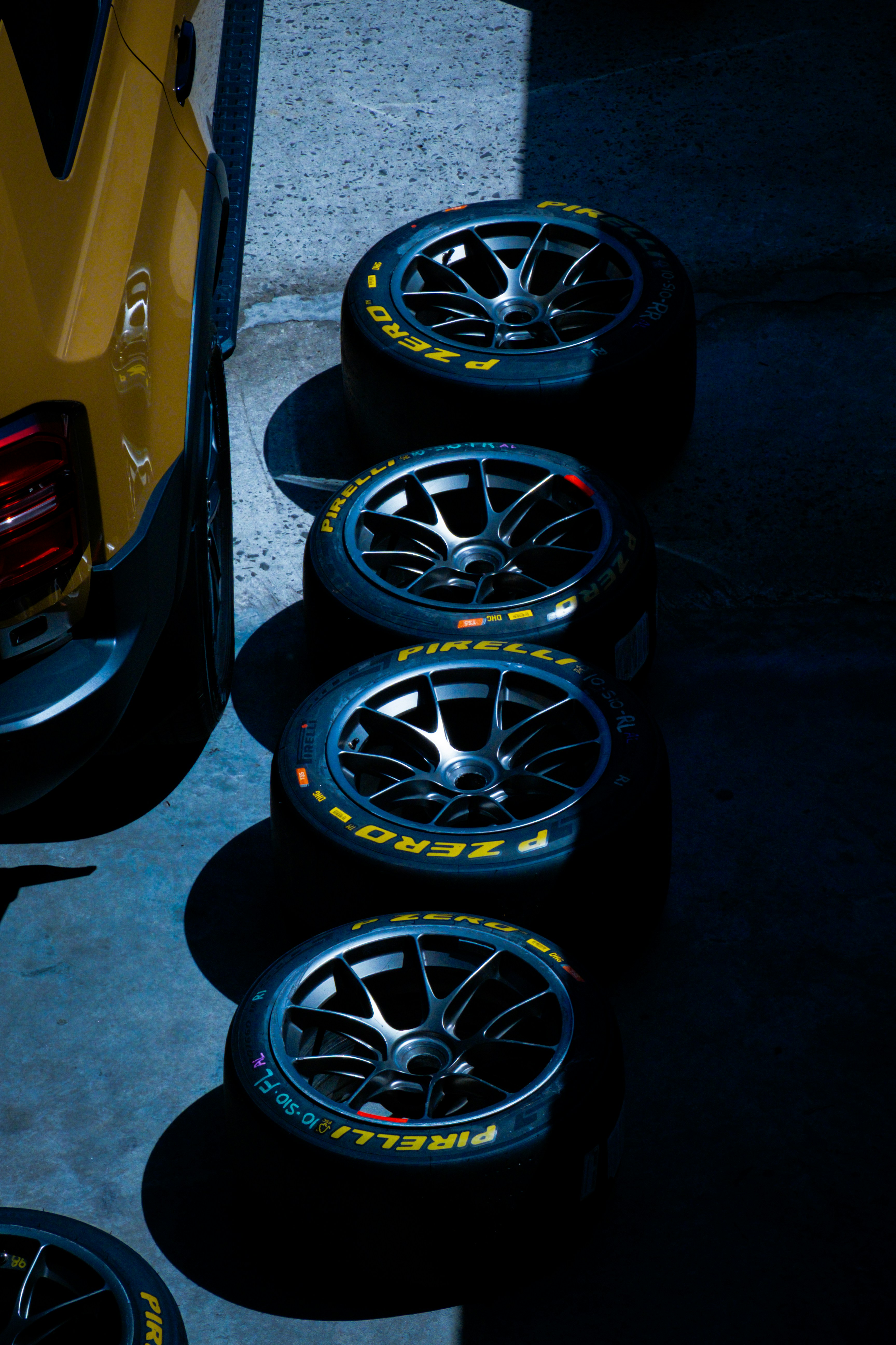 A high-contrast, top-down shot of a row of premium Pirelli P Zero slick racing tyres lined up in the pit lane during the Thailand Super Series at Chang International Circuit, Buriram. The harsh paddock sunlight cuts across the textured concrete, highlighting the bold yellow lettering, aggressive wheel spokes, and smooth rubber surfaces against deep, dramatic shadows. Perfect for motorsport, automotive, and high-performance design projects.
