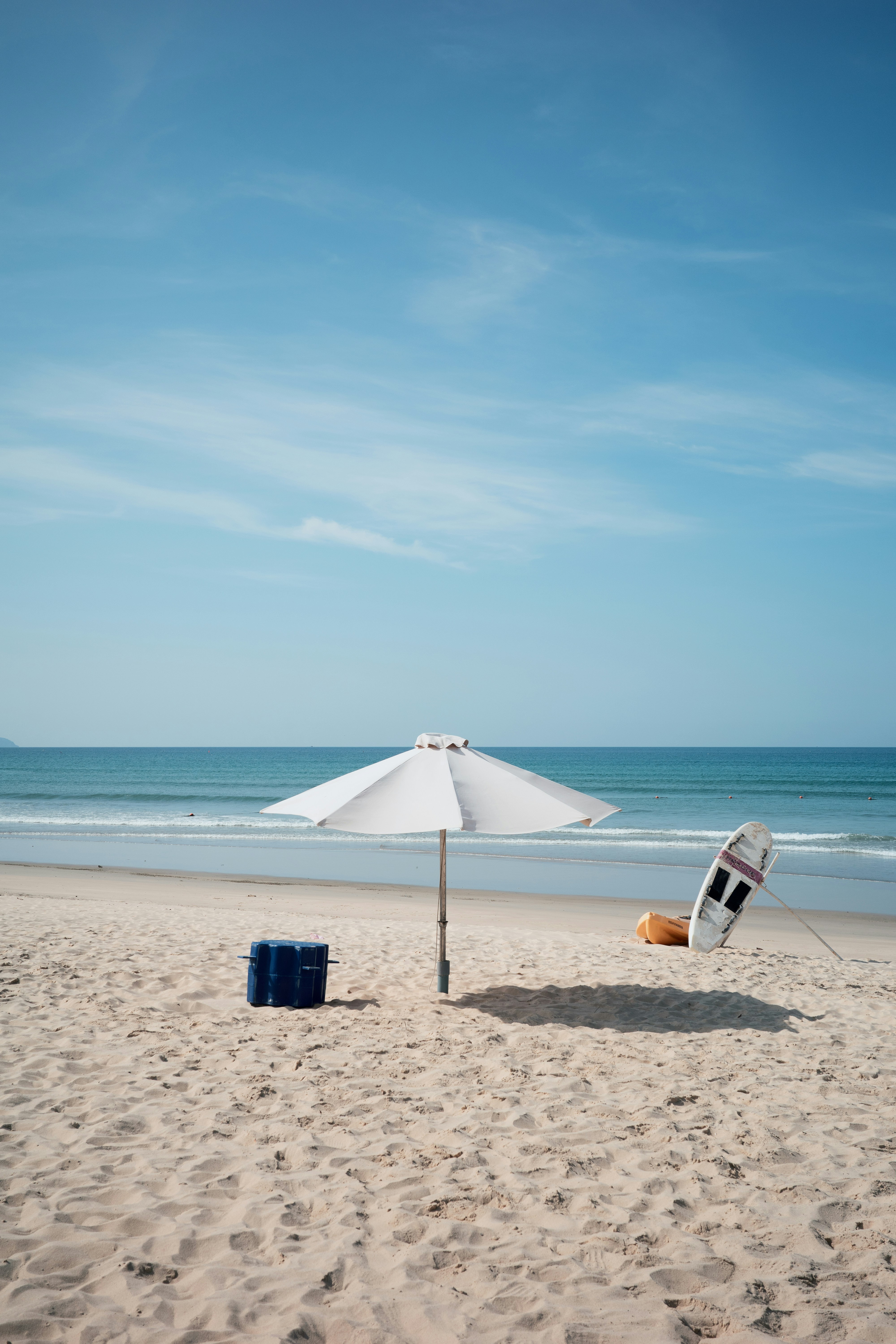 A white umbrella, cooler, and surfboard on a sunny beach.