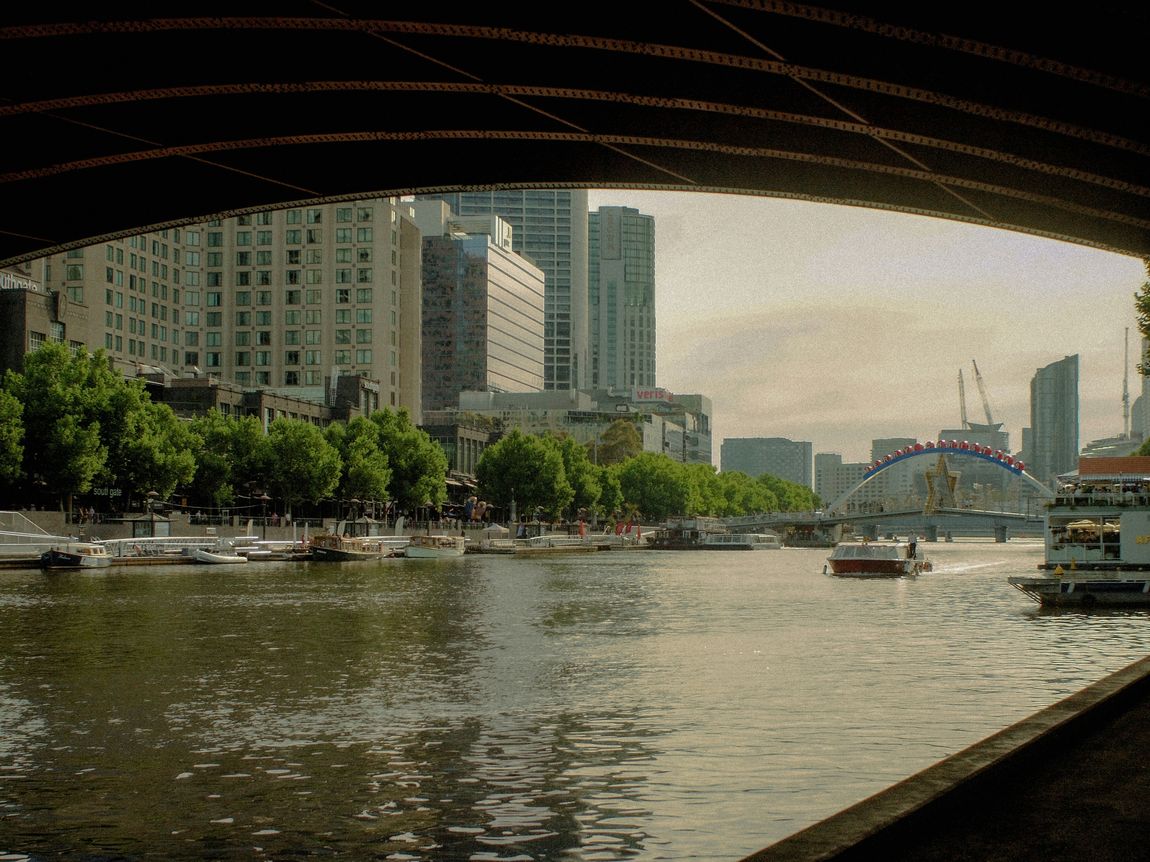 Urban river scene with boats along a city embankment framed by a dark bridge arch; mid-day light highlighting modern buildings and green trees beside the water.