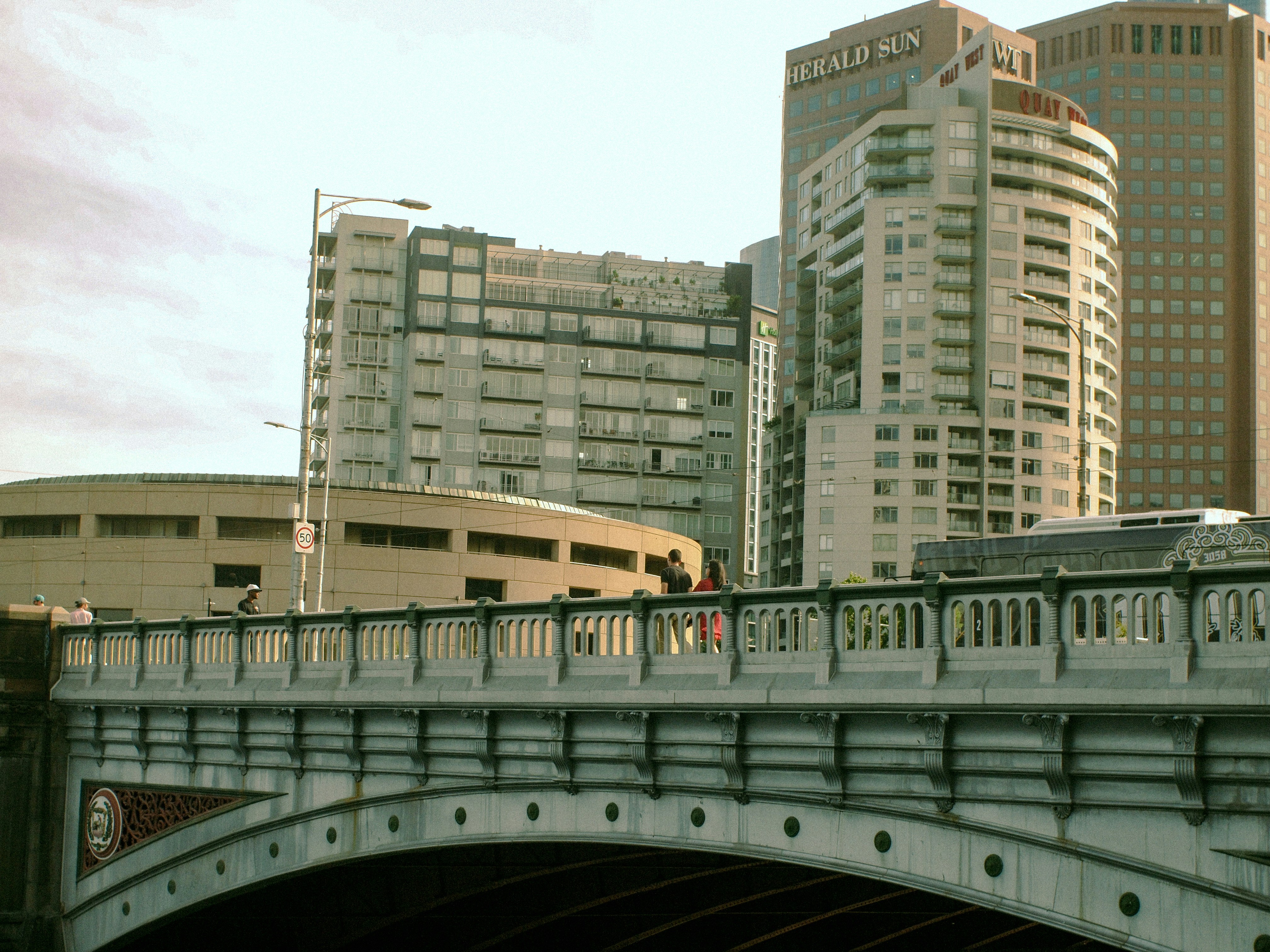 Pedestrian bridge spanning a city canyon with tall office buildings in the background; the scene emphasizes architectural scale and urban rhythm.