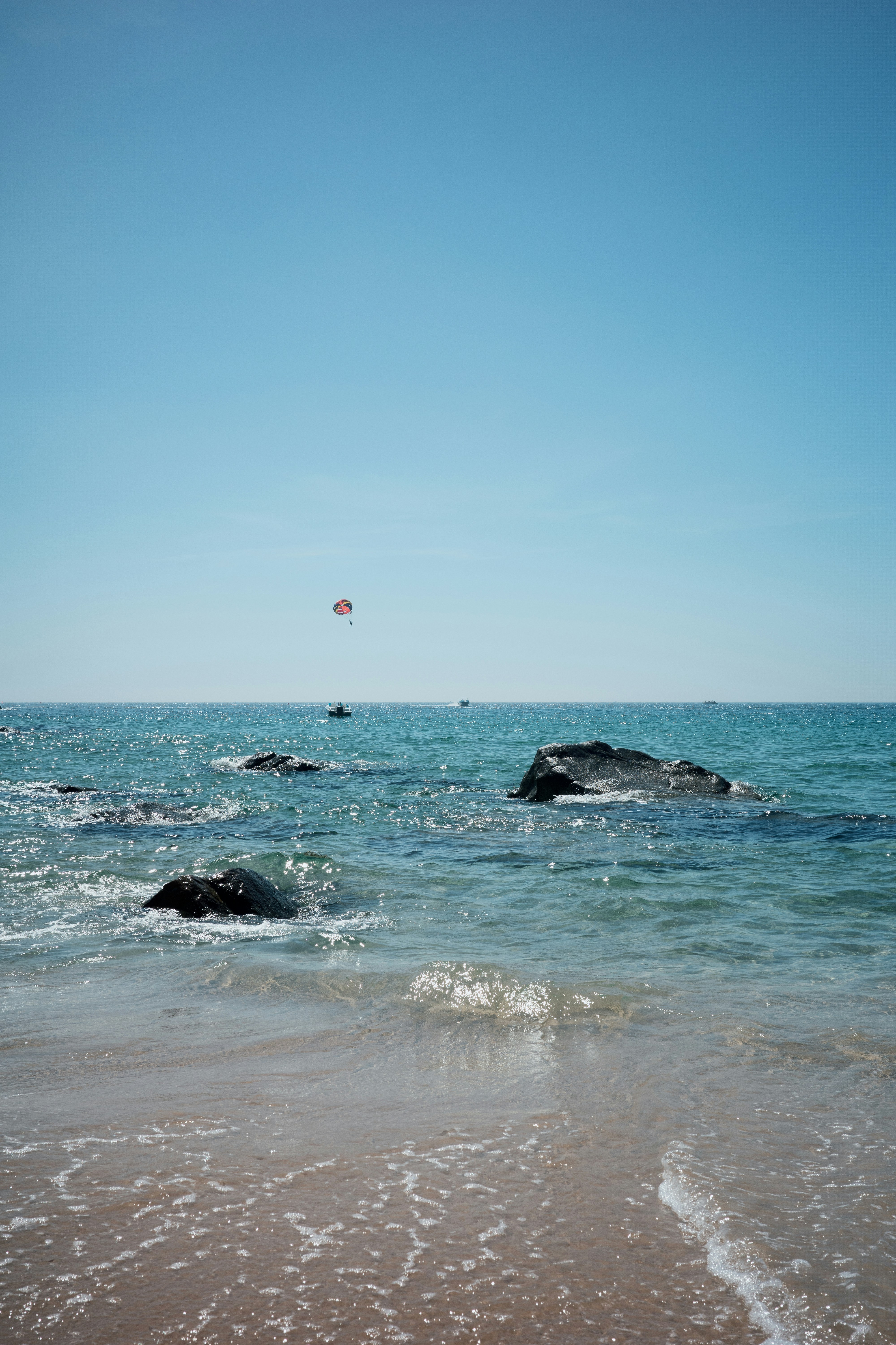 Soft blue sky over a tranquil sea with a distant parachute and small boats; foreground shows gentle waves and rocky outcrops along the shoreline. (Photo is a landscape scene) 