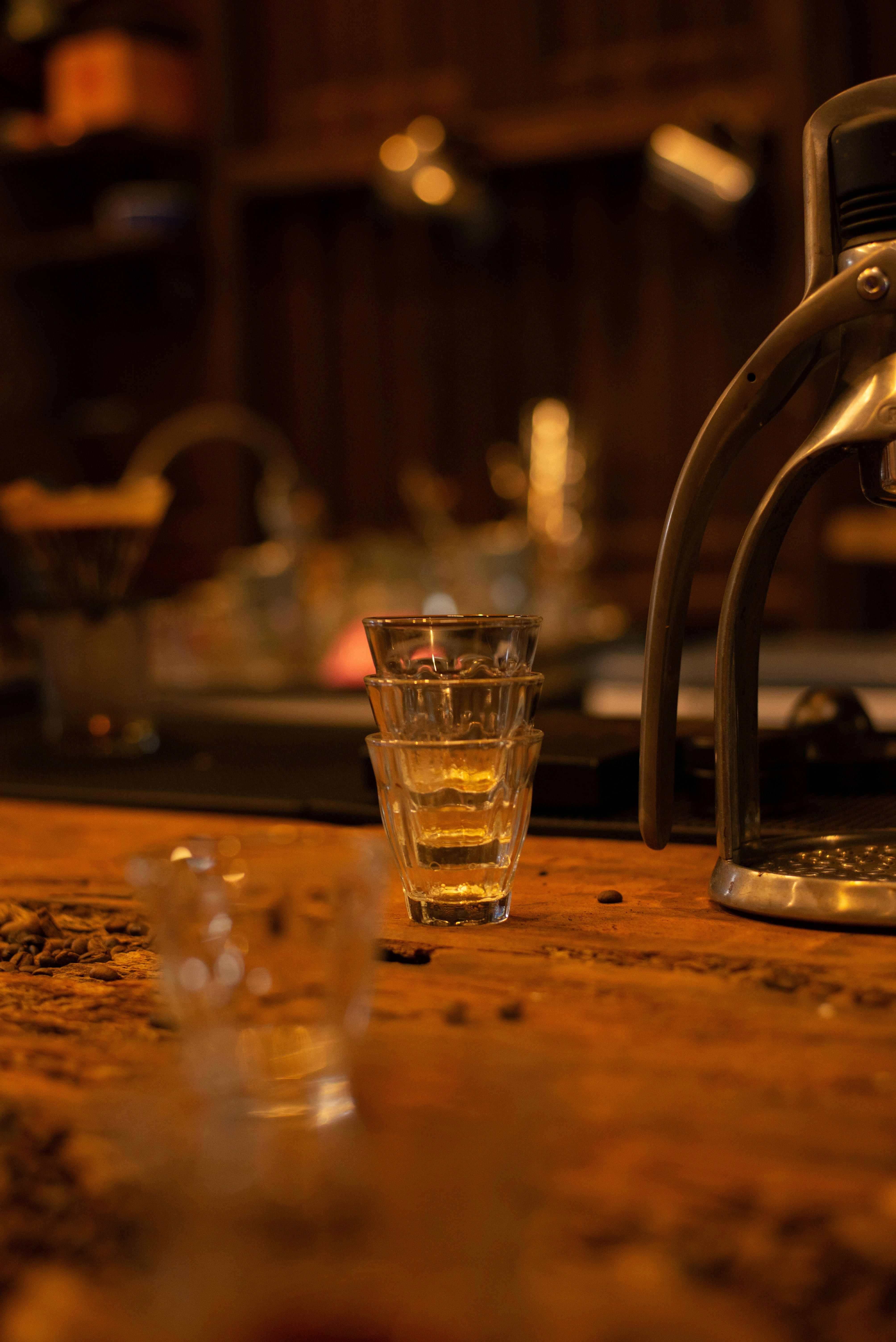 Low-key bar scene with a stack of small glasses reflecting warm light; foreground glass out of focus while a drink and espresso machine sit to the right, creating depth and mood.