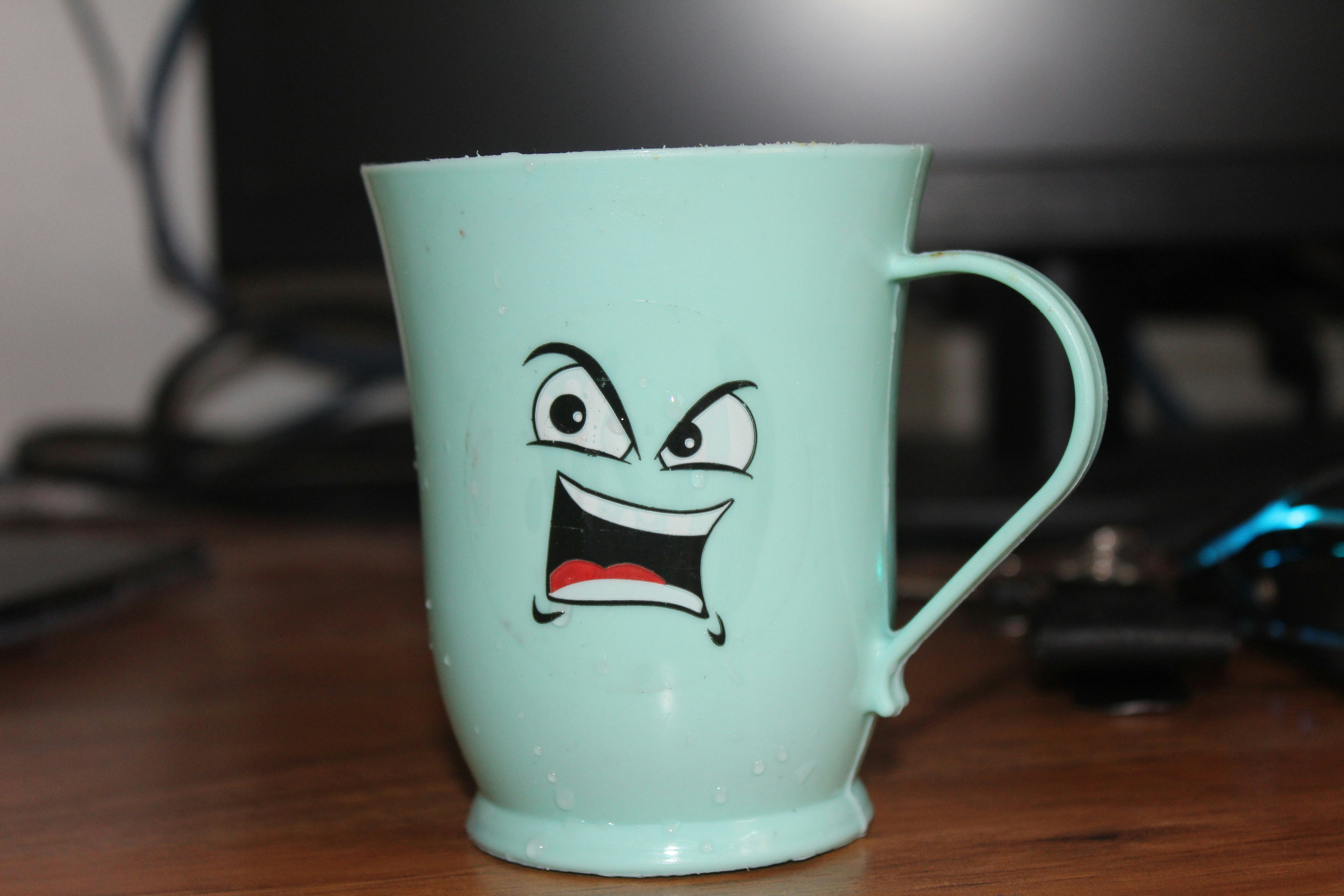 Cartoon-faced teal mug sits on a wooden desk with computer peripherals in the blurred background.