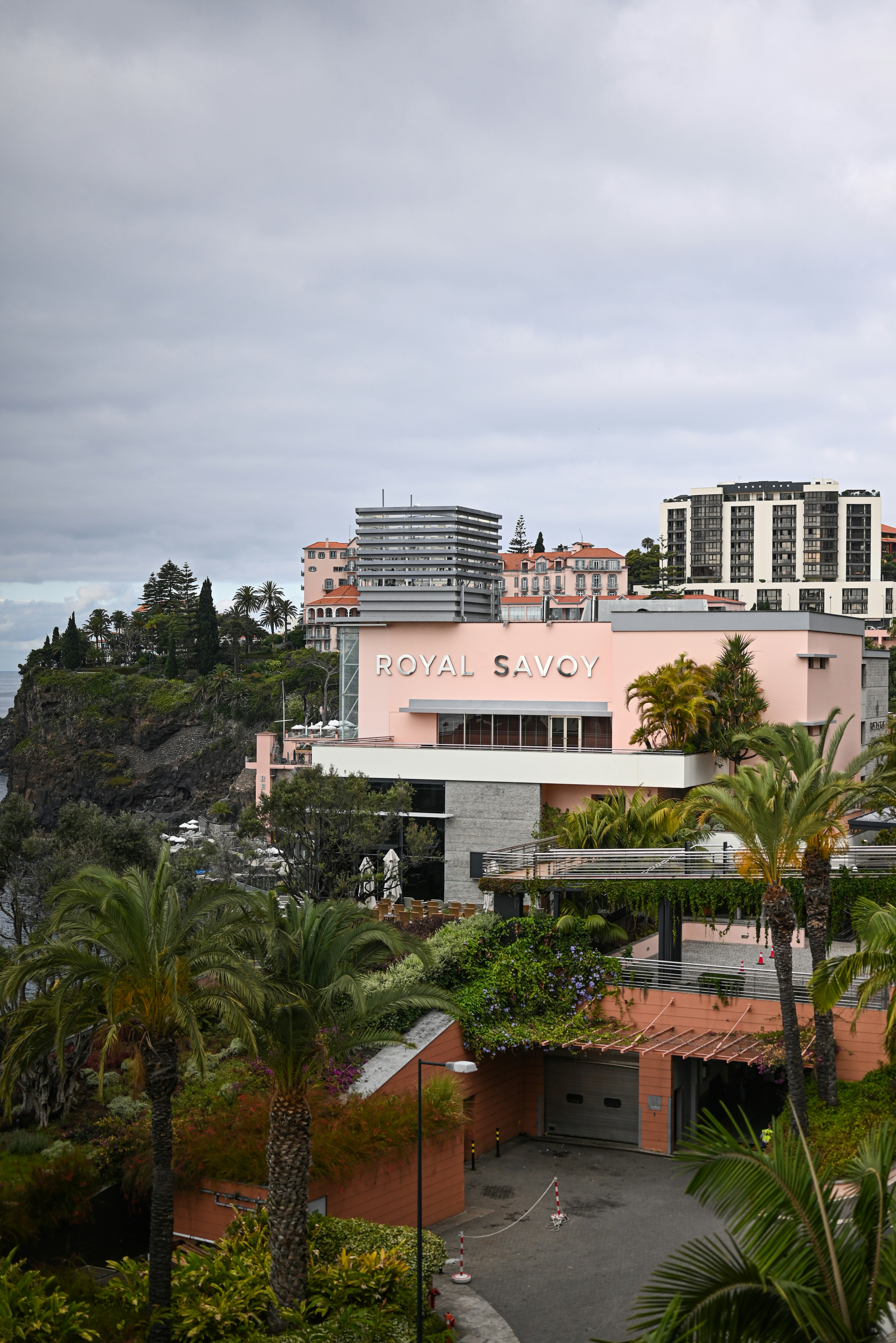 Pink Royal Savoy hotel facade with palm trees and hillside buildings under a cloudy sky, showcasing coastal architecture and greenery.
