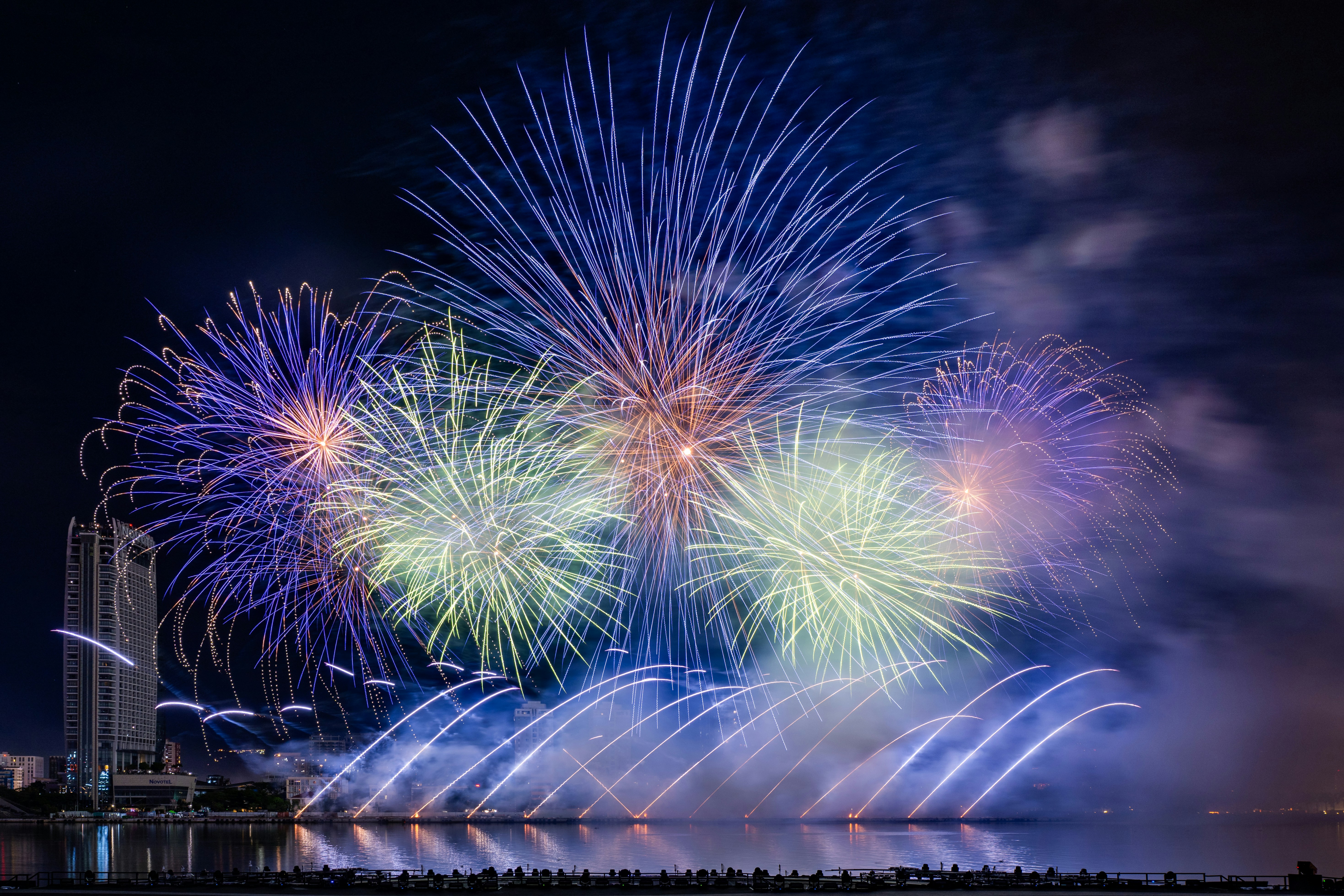 Colorful fireworks explode over a riverfront cityscape, with reflections on the water and light arches skimming the surface.