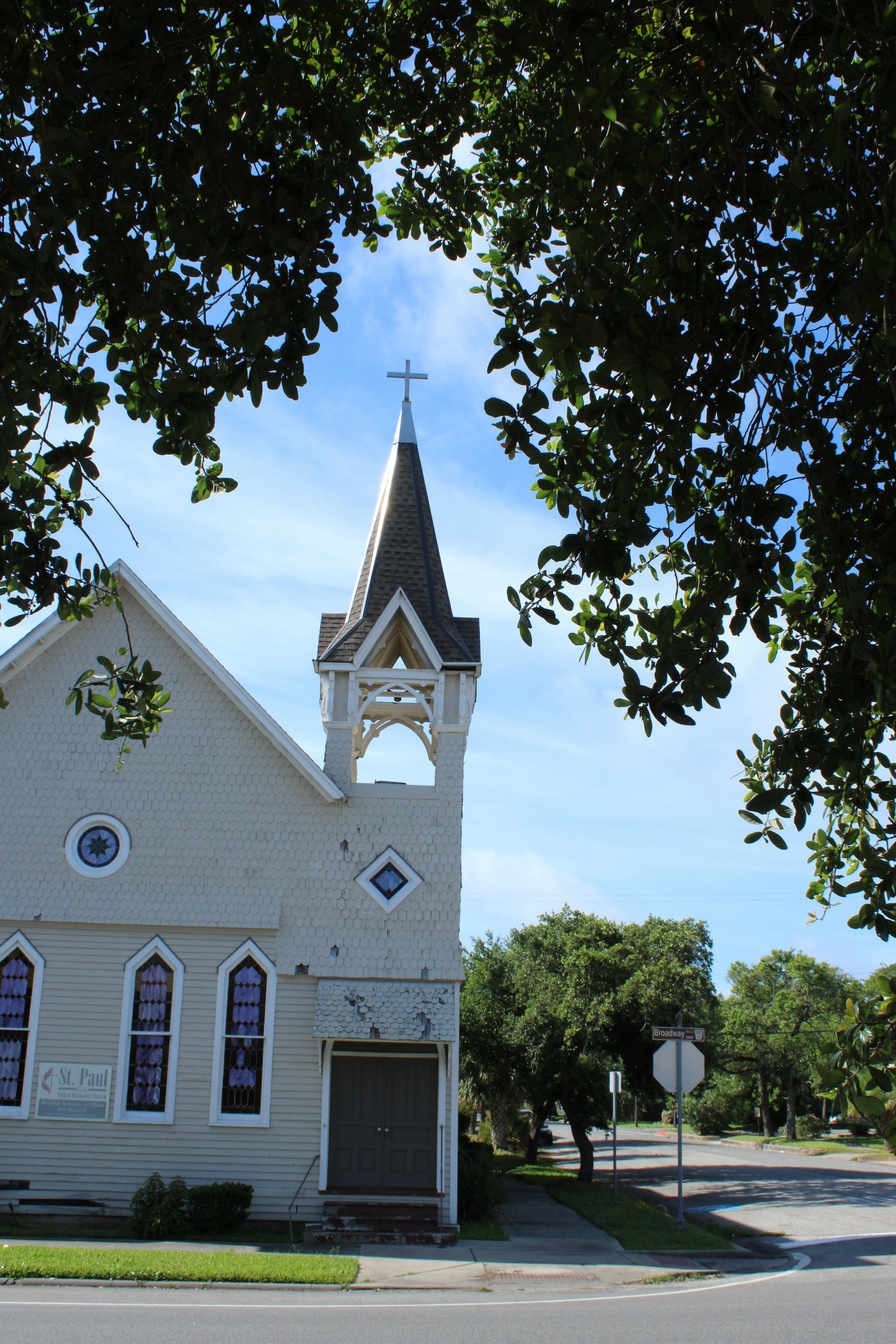 A small white church sits on a corner with a tall steeple reaching toward a blue sky, framed by overhanging tree branches. The scene conveys a quiet neighborhood setting with a street and foliage.