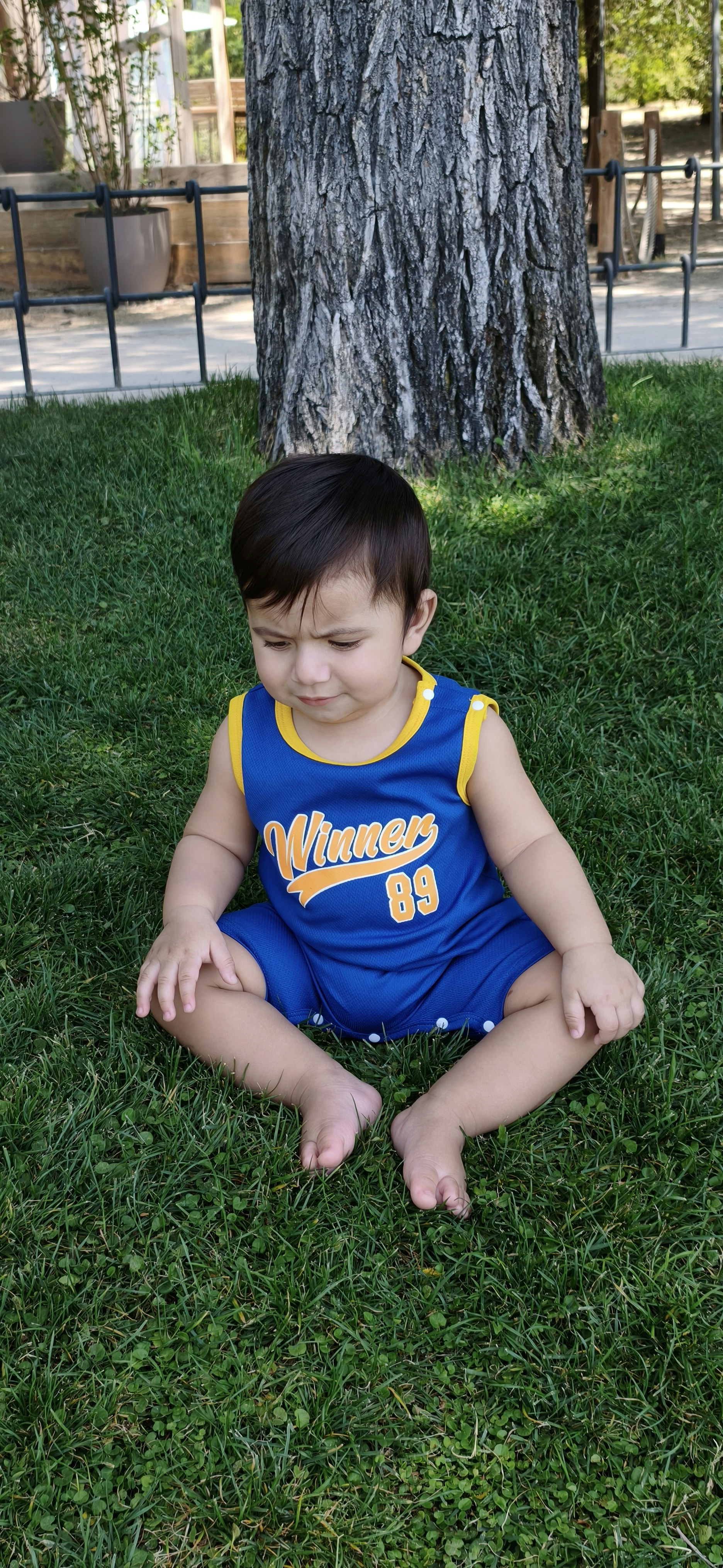 Baby in blue 'winner 89' romper sitting on green grass.