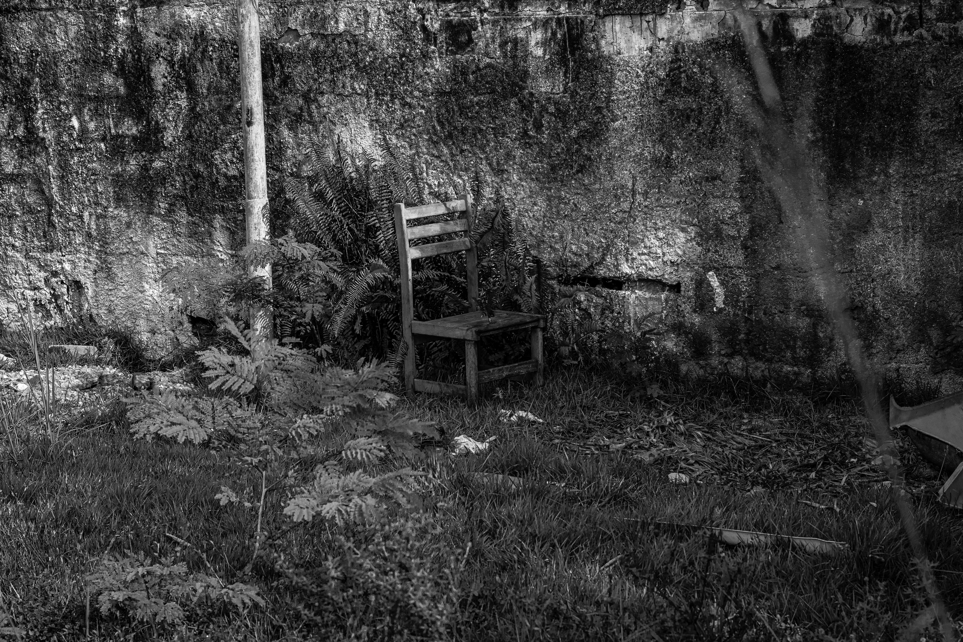 An empty wooden chair sits abandoned against a weathered, moss-covered wall, evoking a sense of loneliness and forgotten memories.