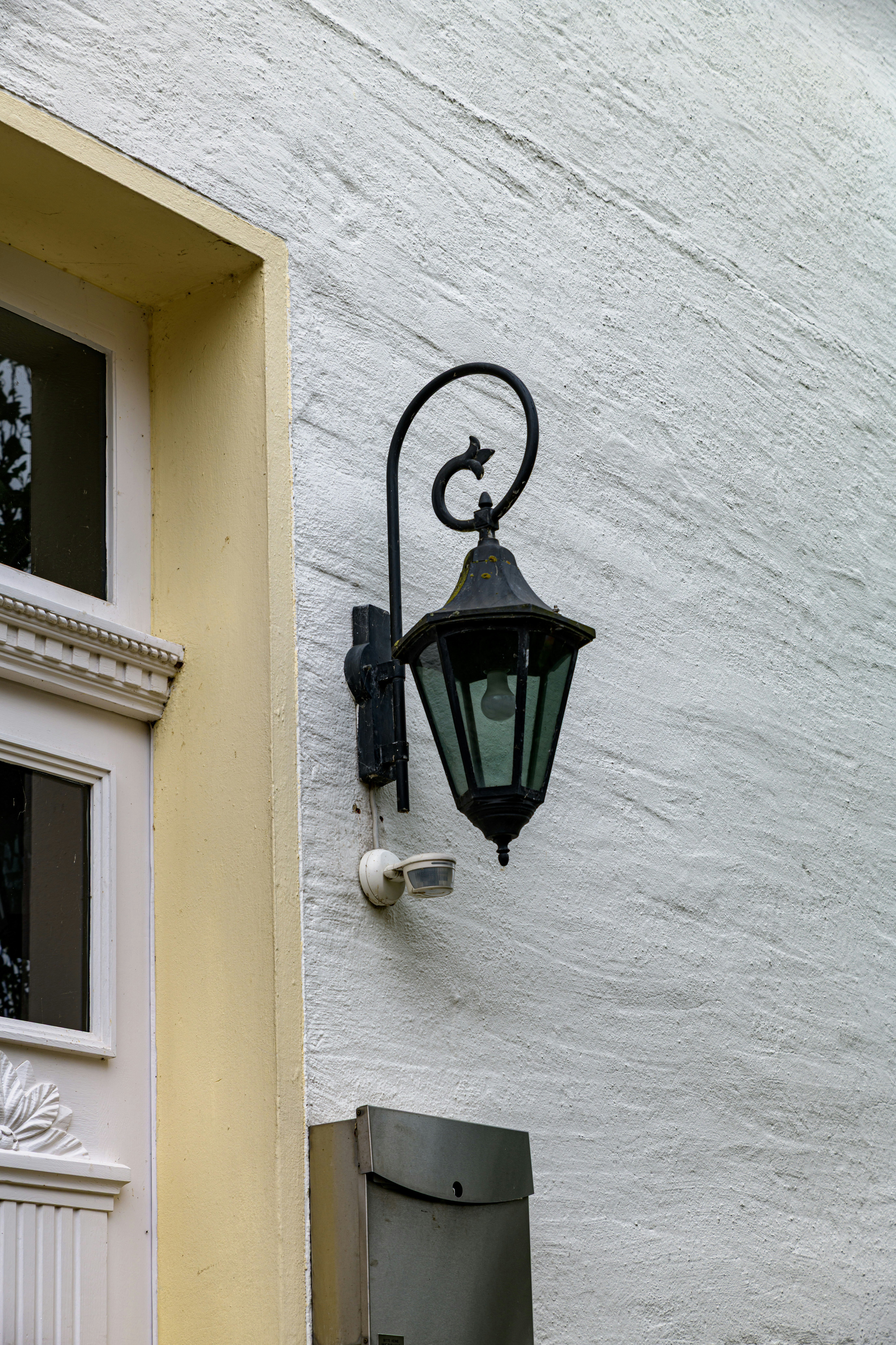 A classic black lantern mounted on a white stucco wall beside a pale-yellow-framed doorway, with a small security camera below. The composition emphasizes the lantern as the focal point of the textured exterior wall.
