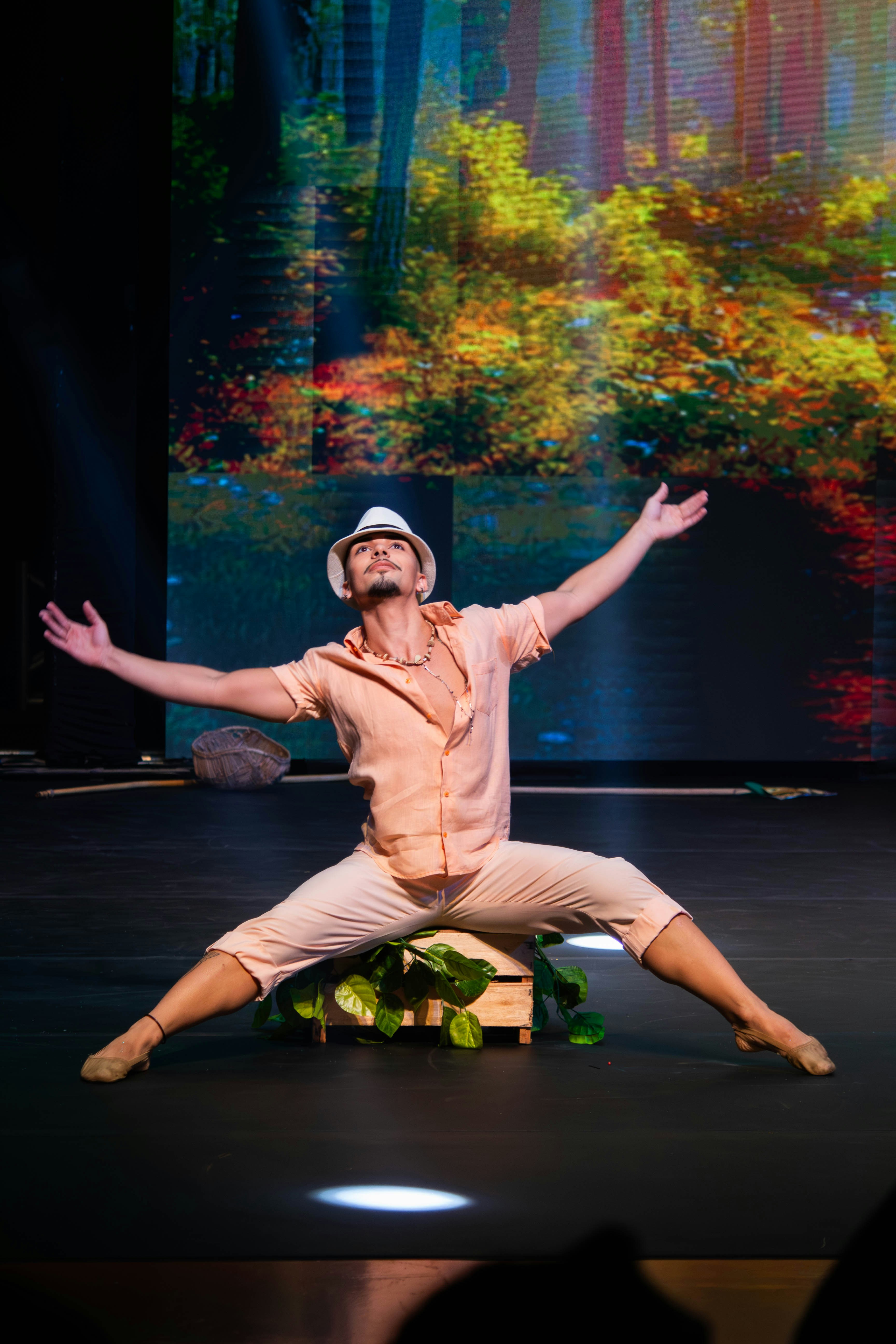 Athletic performer in peach outfit and hat strikes a wide, dramatic pose on stage with a colorful forest backdrop.