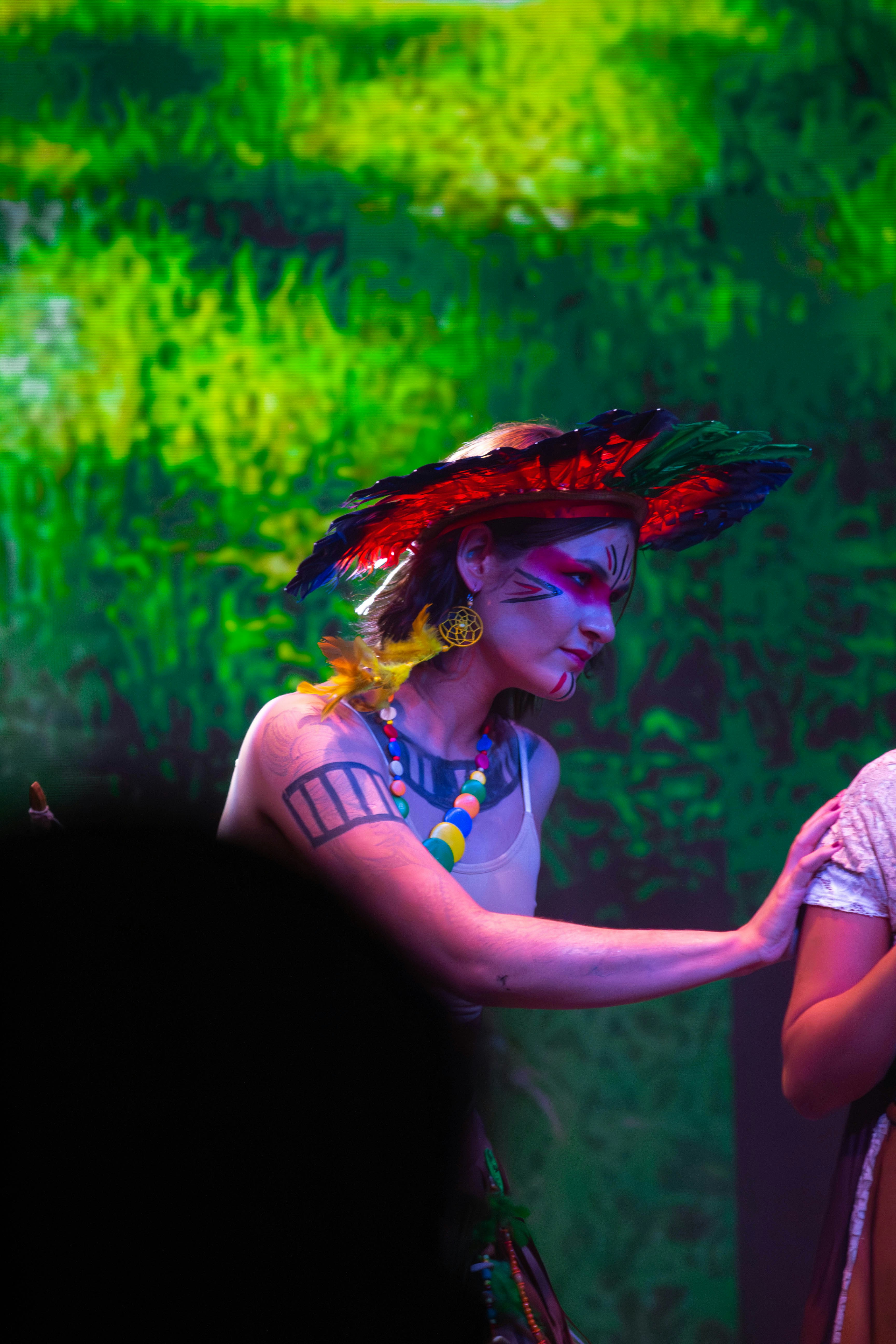 Energetic performer with bold face paint and feathered headpiece engages a companion on a colorful stage.