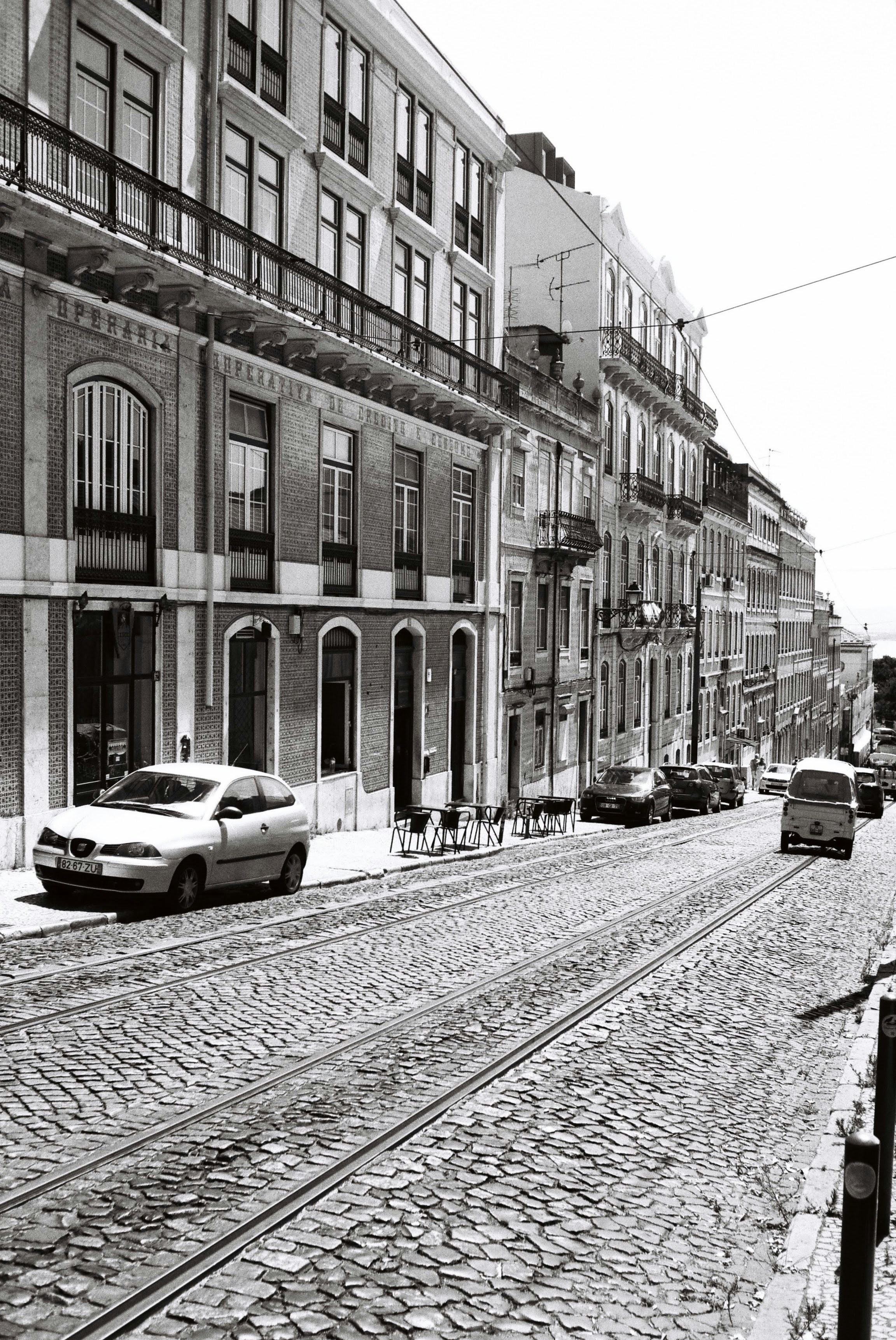 A black-and-white street scene on a cobblestone incline with tram tracks, classic brick buildings, and parked cars lining the curb.