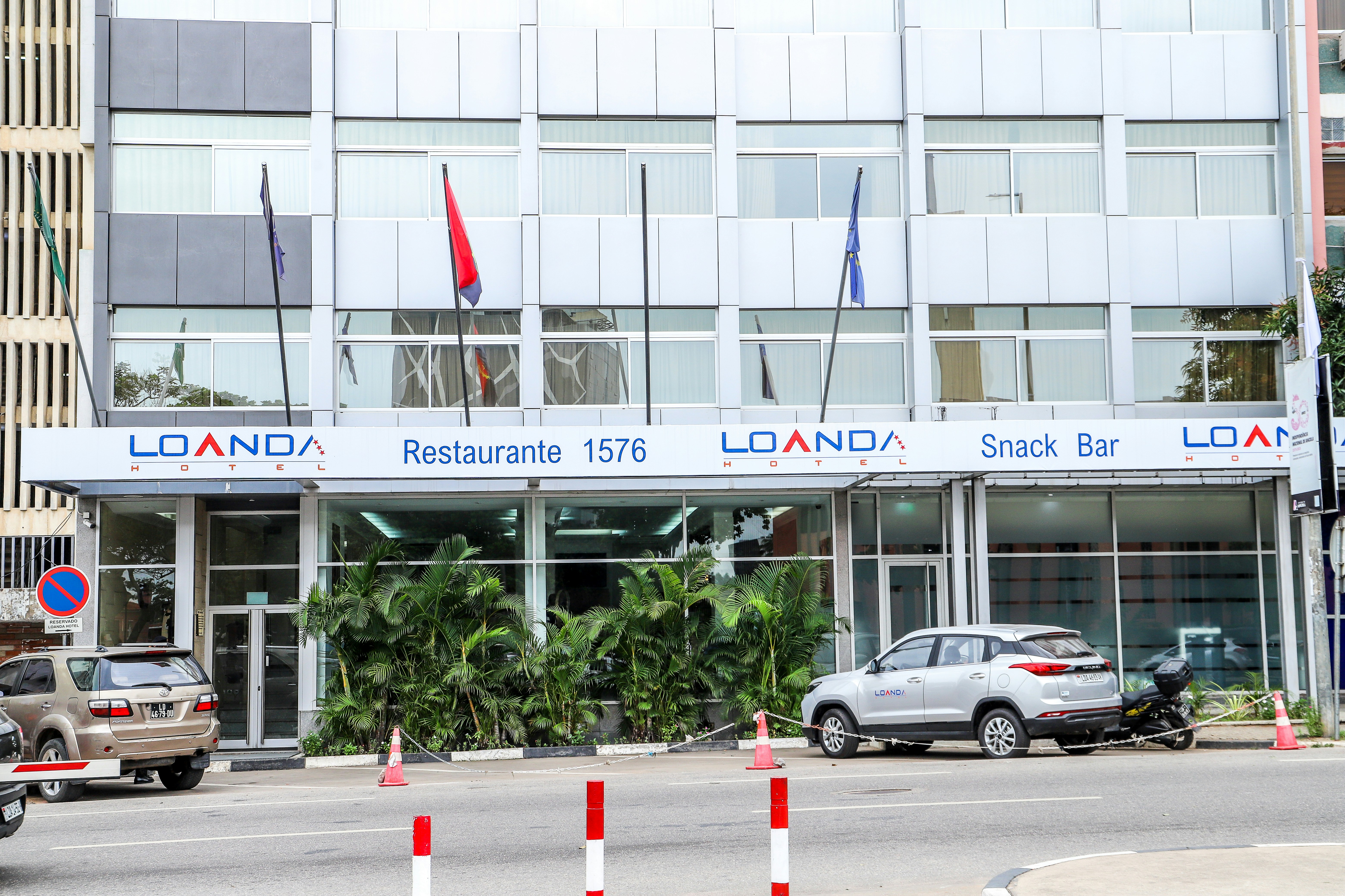 Modern storefront with a glass-front restaurant, signage, and flag poles; vehicles parked along the curb with landscaping in front. Logoed signage indicates a dining establishment.