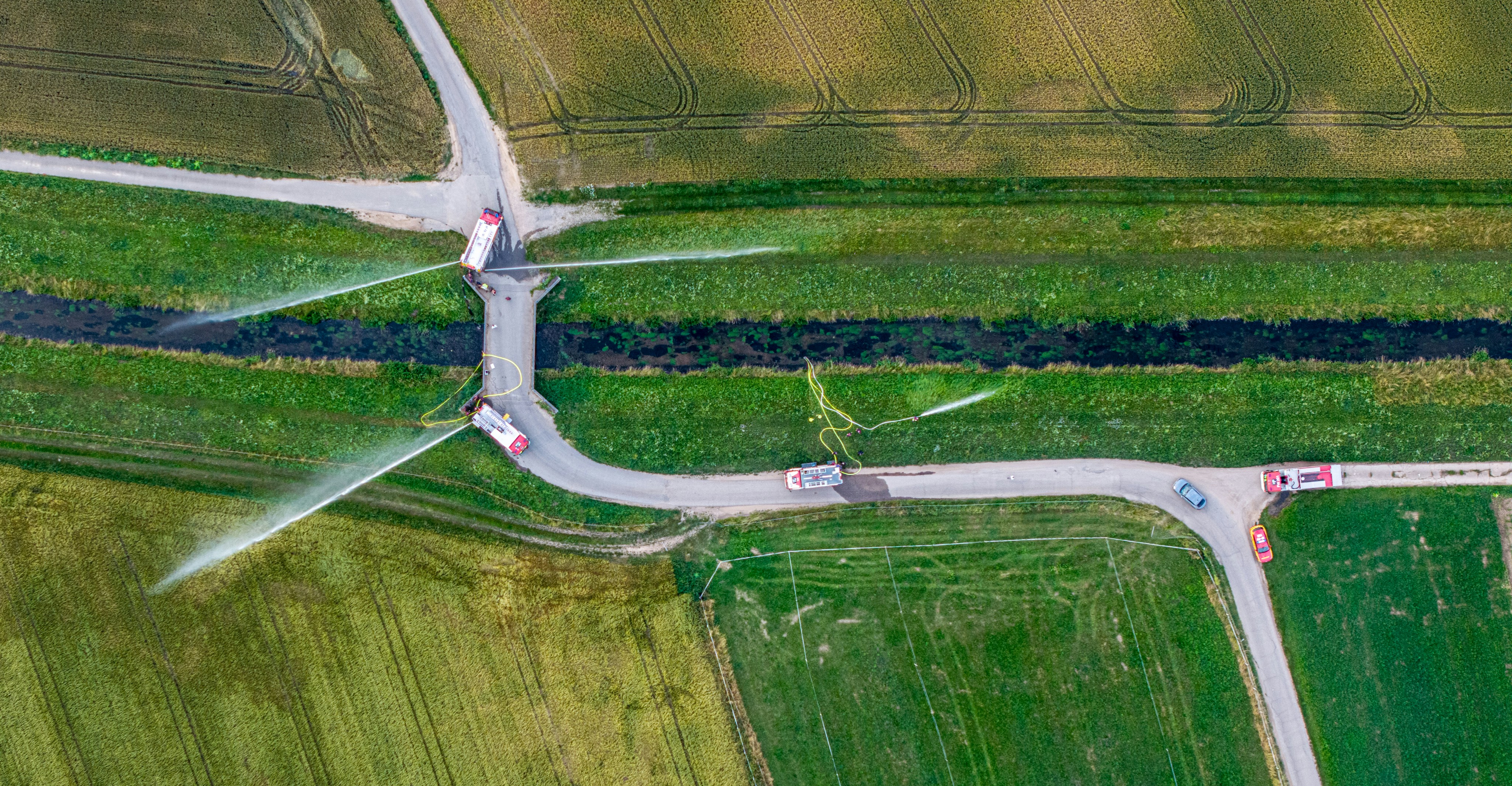 Drone view of a winding rural road crossing a wind turbine and green fields, with vehicles visible along the split path. Aerial landscape capture highlighting infrastructure and agriculture.