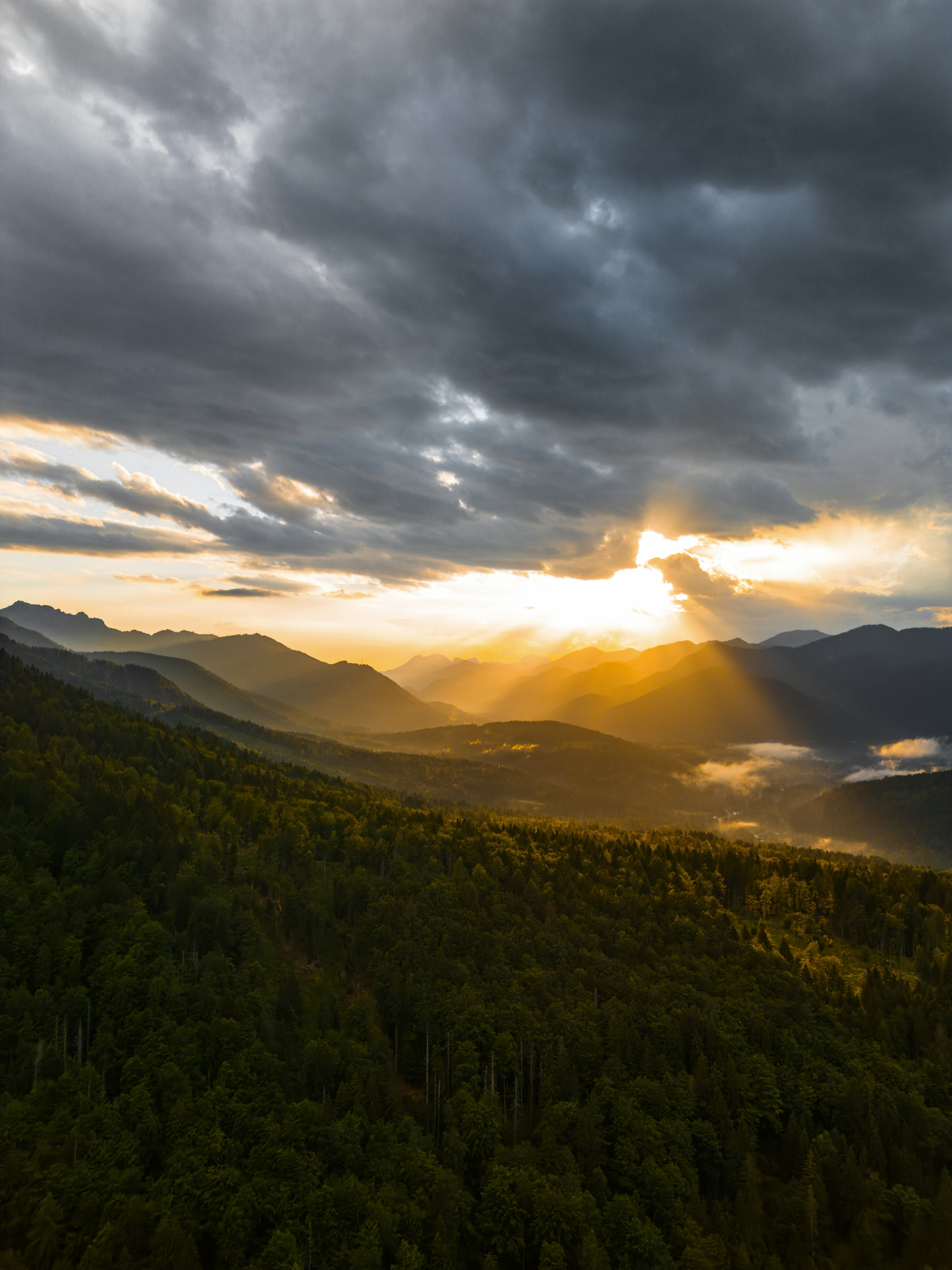 Sunset over forested mountains with dramatic clouds and sun rays.