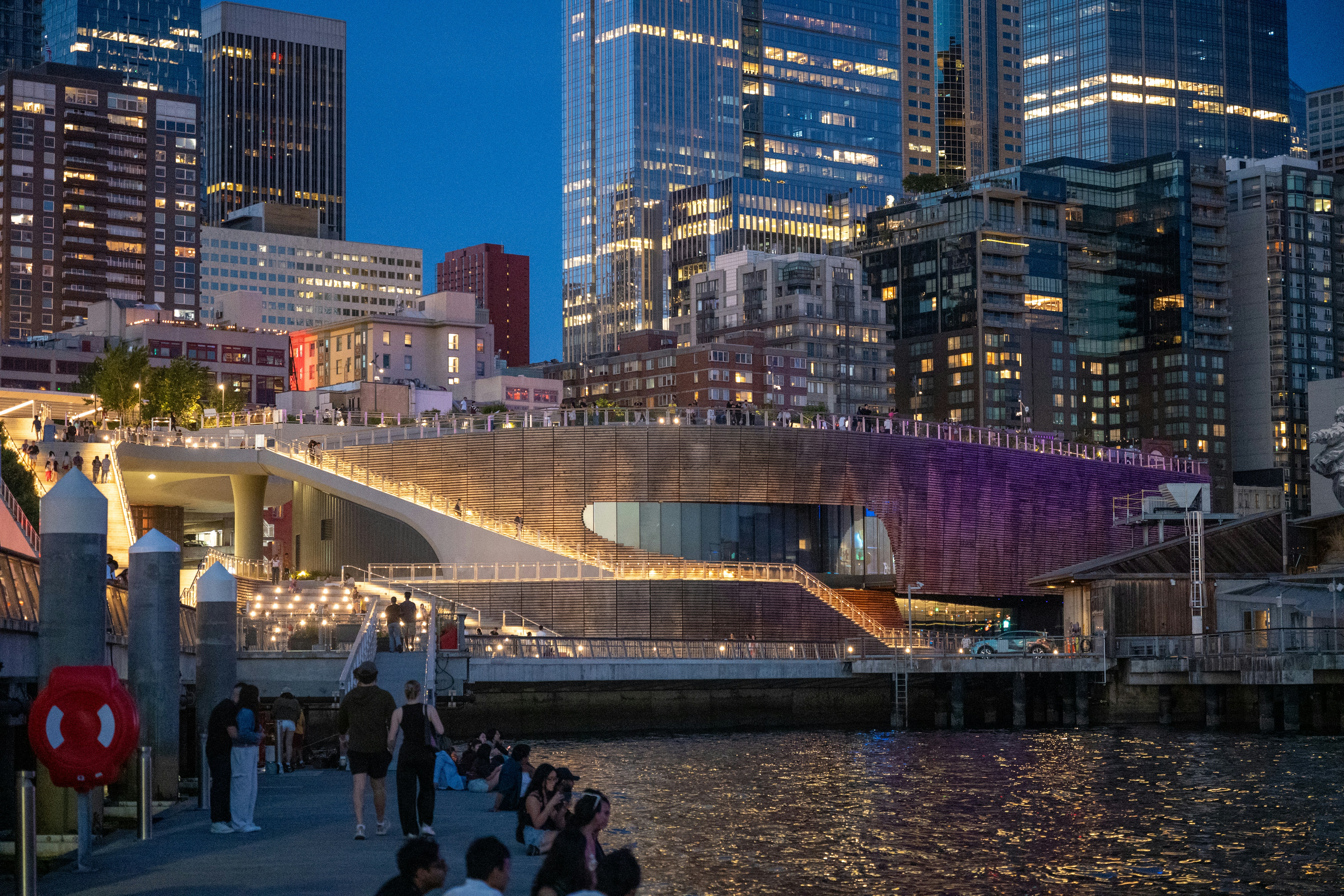 Modern city waterfront with illuminated buildings and people at dusk.